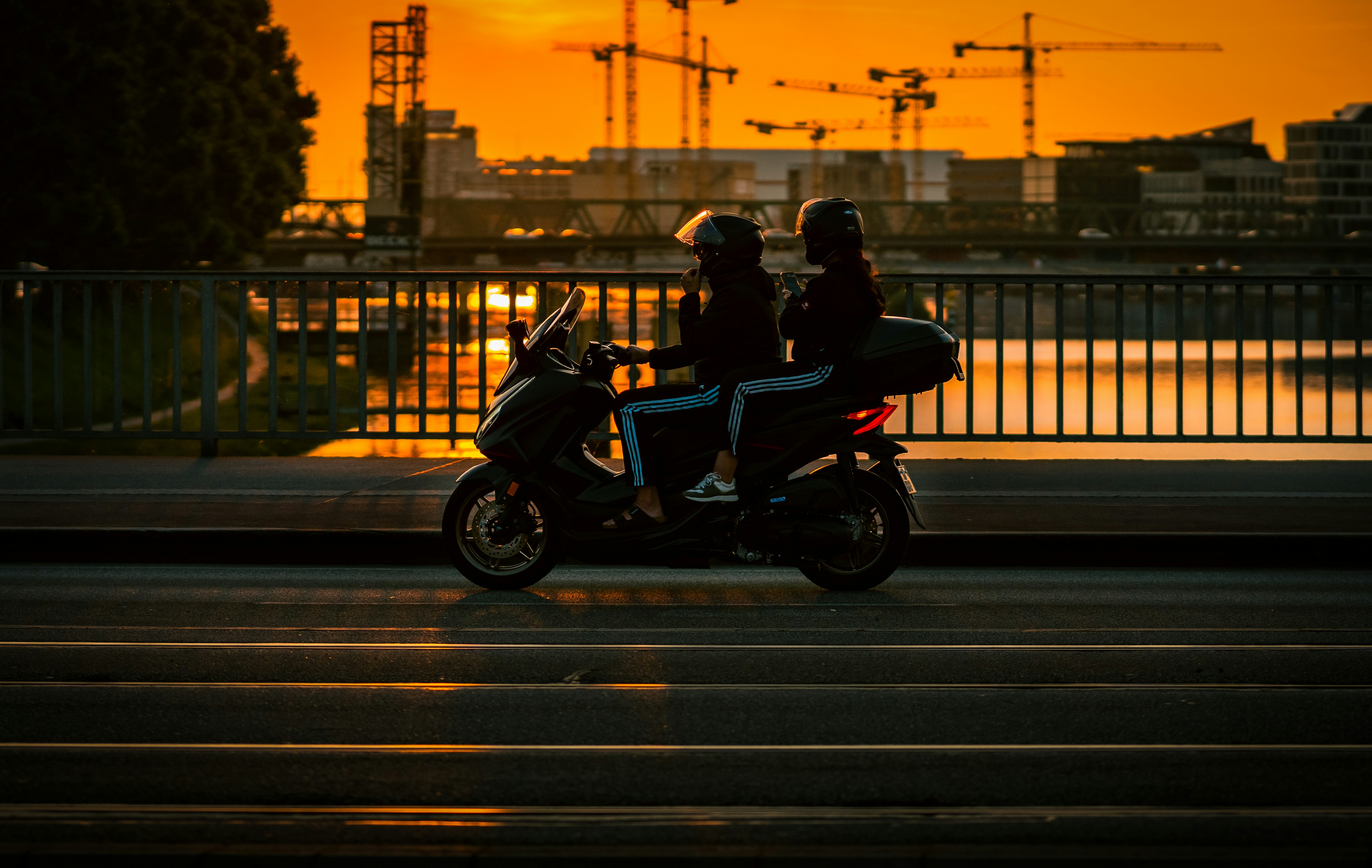 Two riders on a scooter silhouetted against a vibrant sunset, with construction cranes in the background reflecting the day's end.