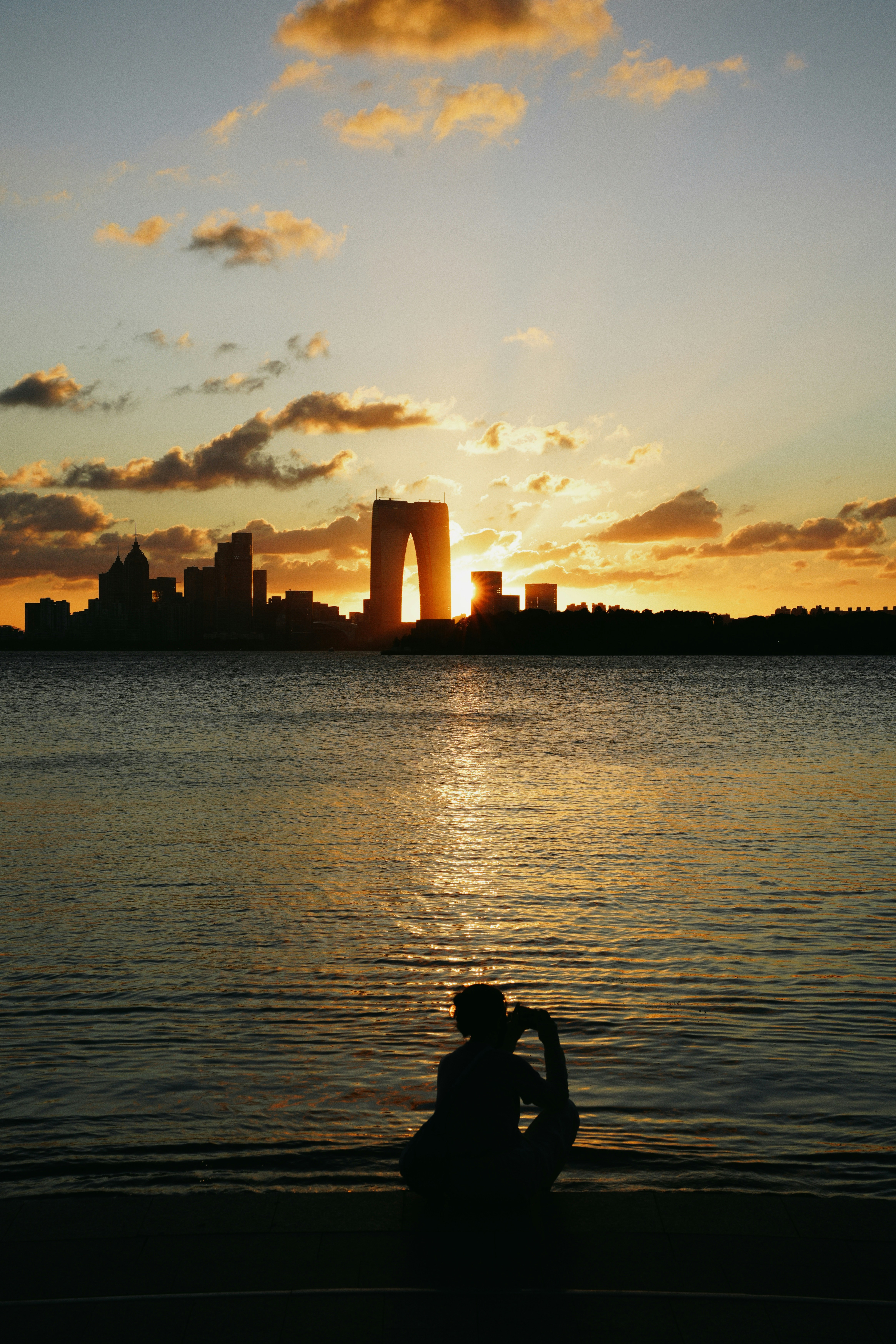 Silhouette of a person sitting by the water, framed against a vibrant sunset with city skyline in the background.