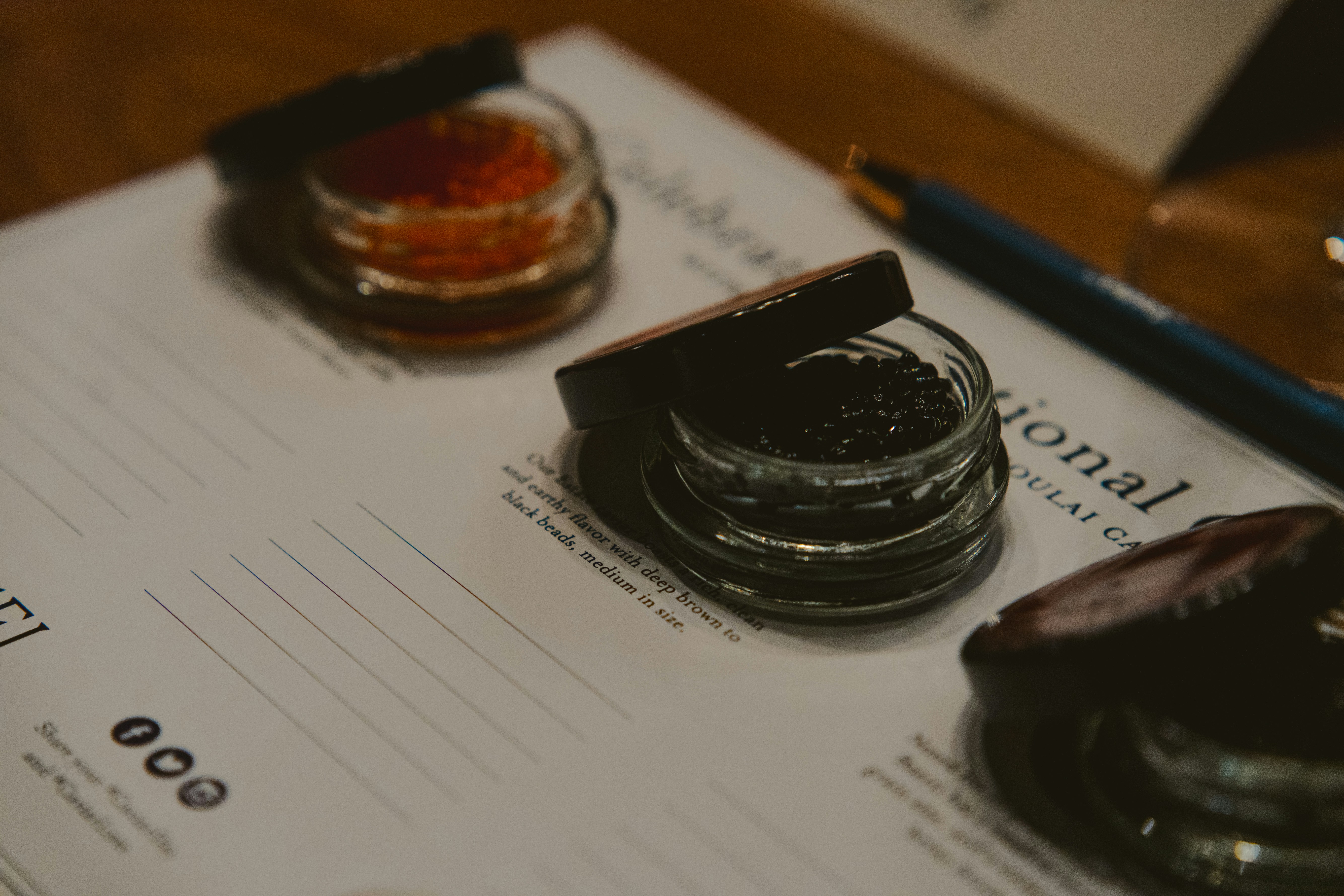 Three small glass jars filled with vibrant condiments rest on a menu, showcasing a blend of colors and textures. A pen lies nearby, hinting at a culinary experience.