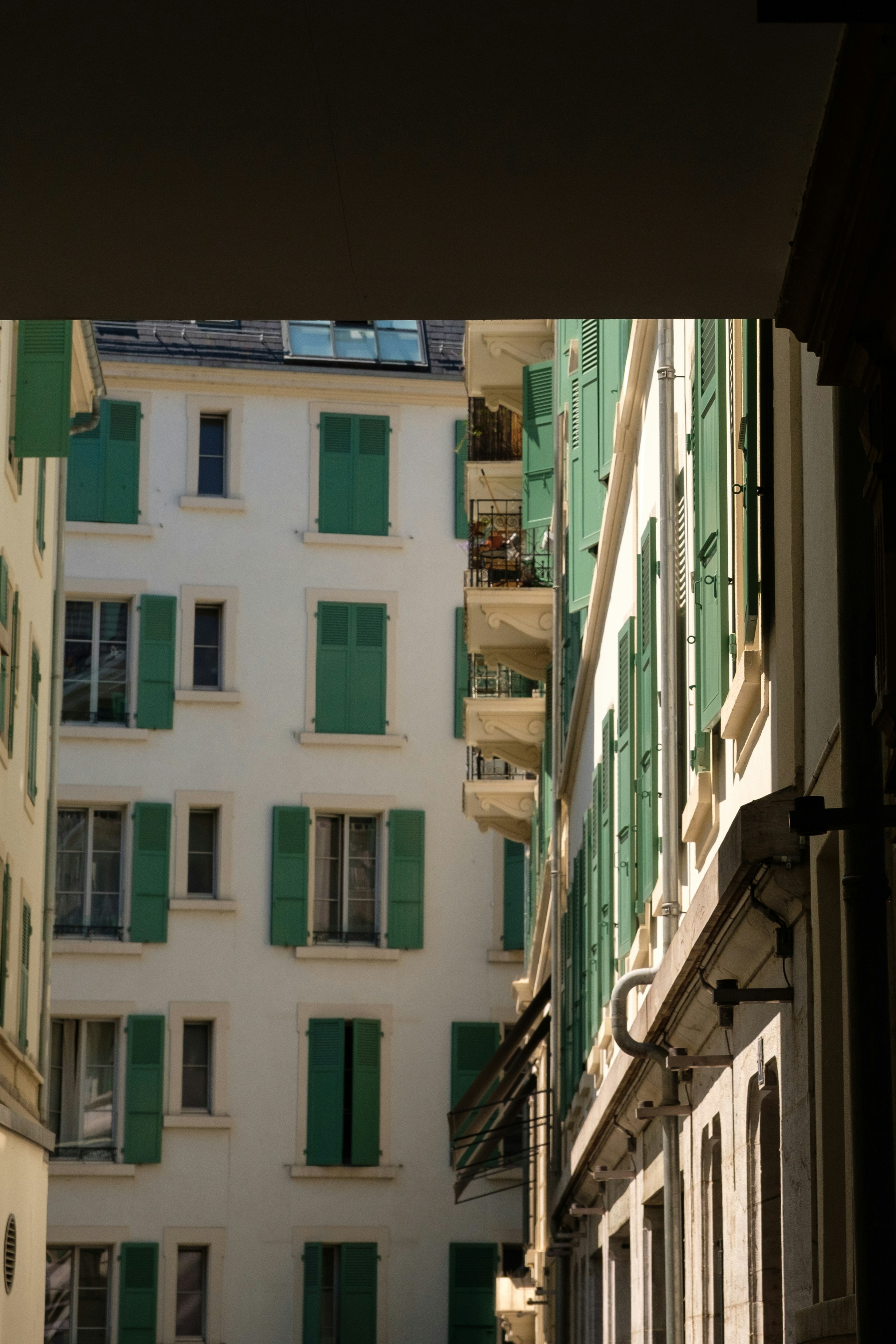 Sunlit alleyway revealing a glimpse of buildings adorned with vibrant green shutters and balconies.