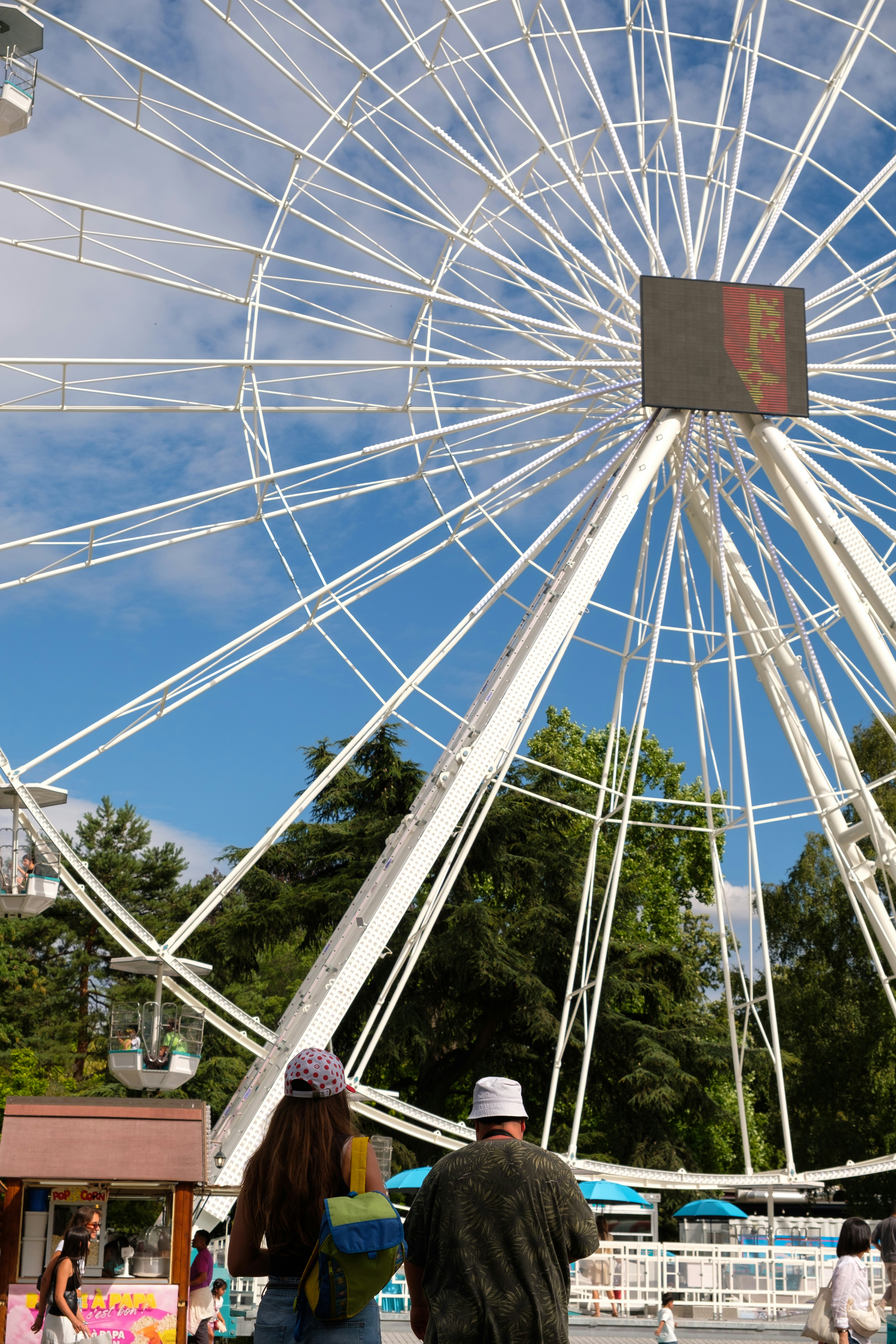 Two visitors stand in awe beneath a towering Ferris wheel, framed by a vibrant sky and lush greenery.