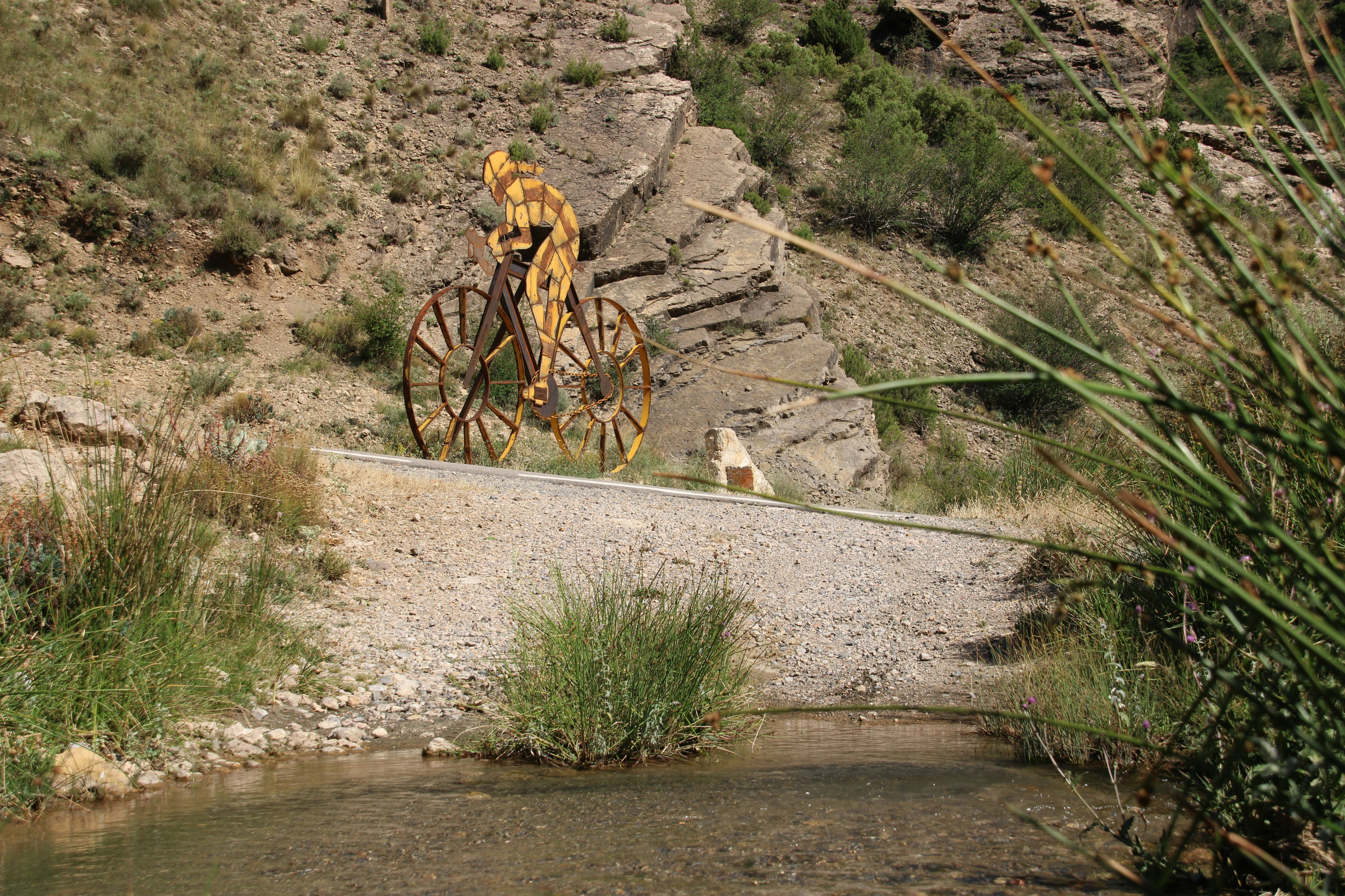 Hilltop Cyclist: sunlit metal sculpture perched above a rugged gravel trail