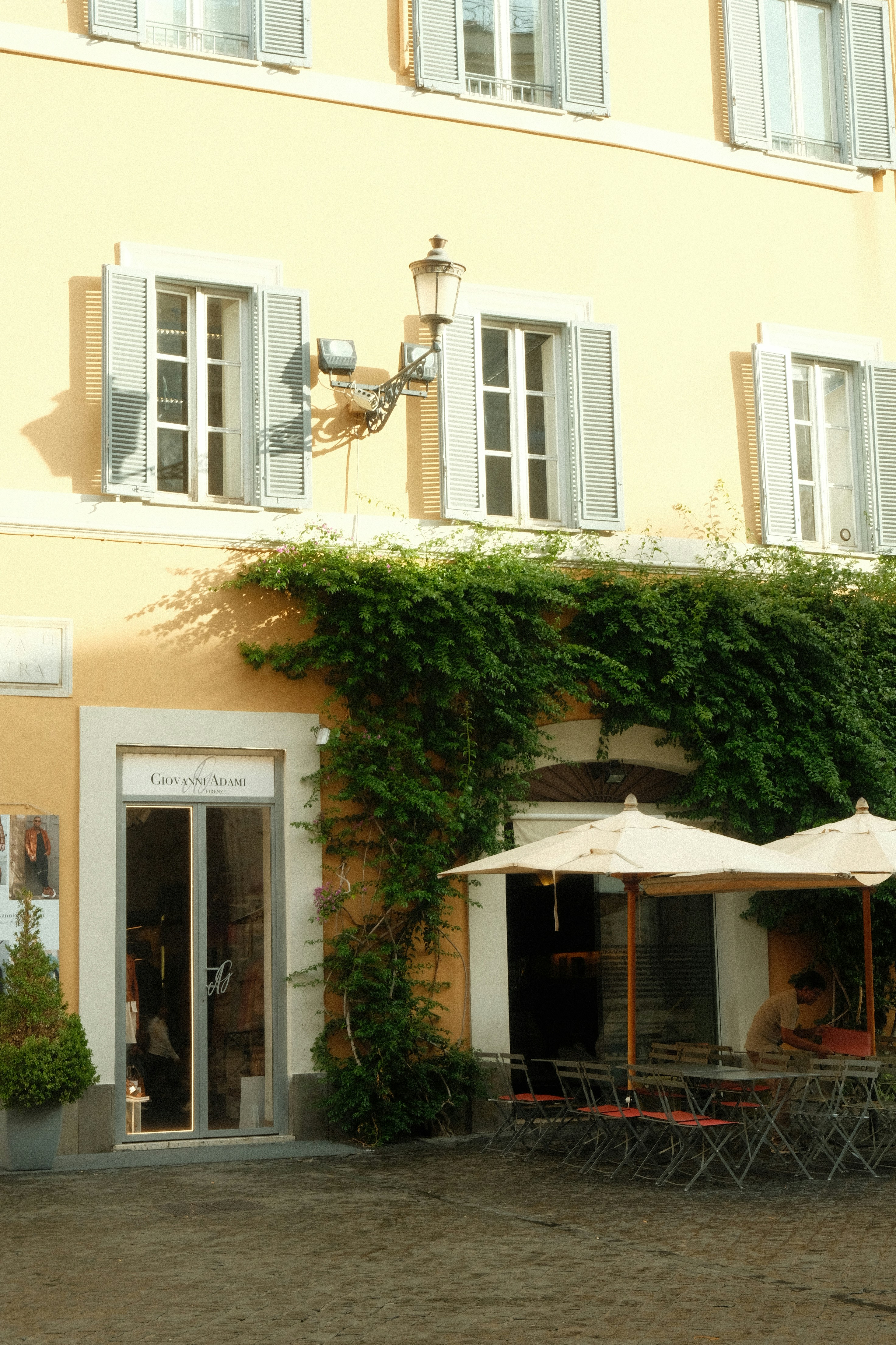 Quaint café entrance adorned with lush greenery, inviting patrons to enjoy a serene moment. Sunlit façade enhances the warm ambiance.