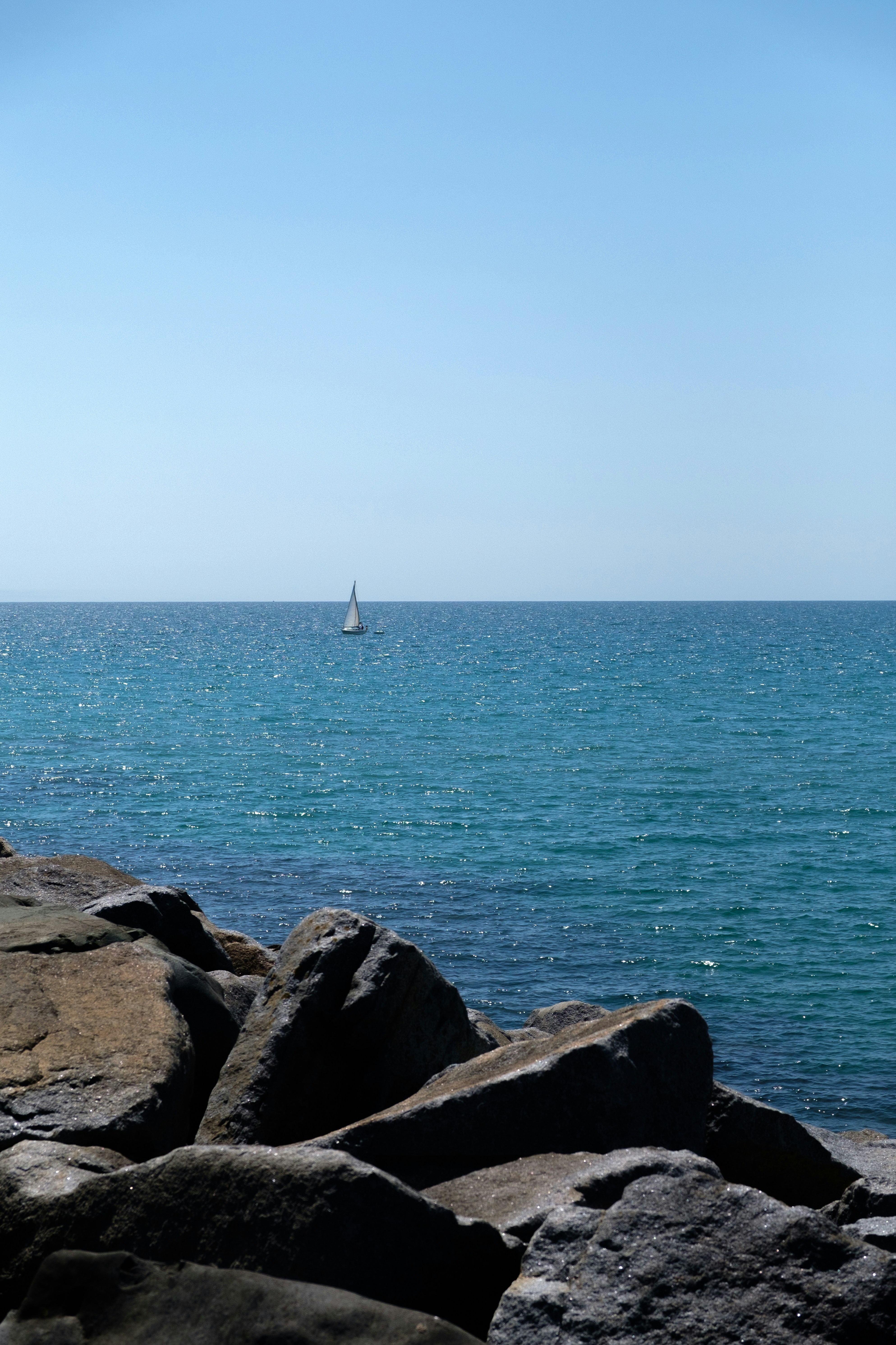 A solitary sailboat glides across a shimmering sea, framed by rugged rocks in the foreground. The vast blue sky and water create a tranquil atmosphere.