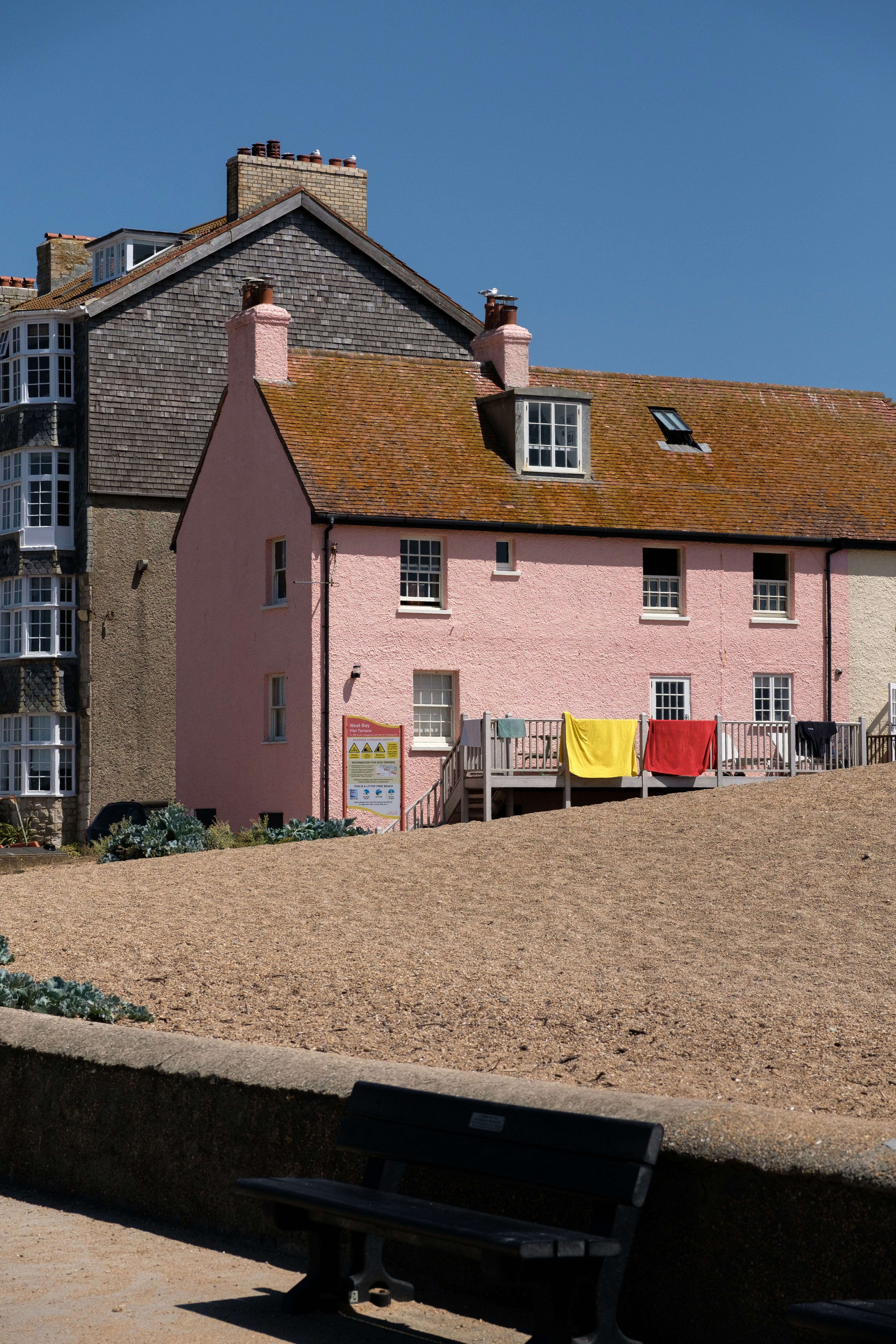 A pastel pink house with a sloping roof stands beside a grey building, adorned with colorful laundry hanging on the porch. The scene captures the essence of seaside living.