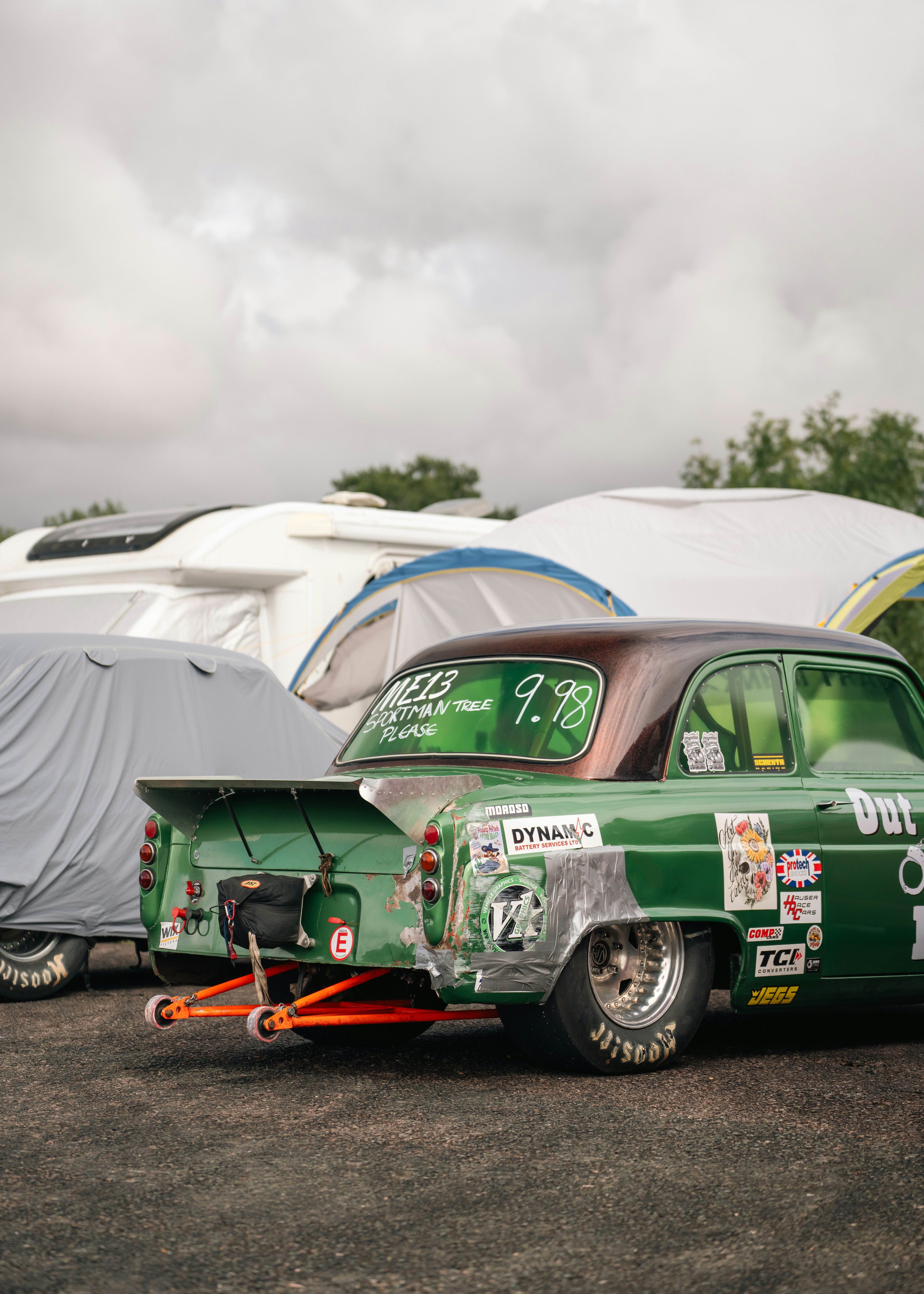 Modified green classic car with racing decals parked beside covered vehicles at a racing event. The car showcases a unique design and performance modifications.