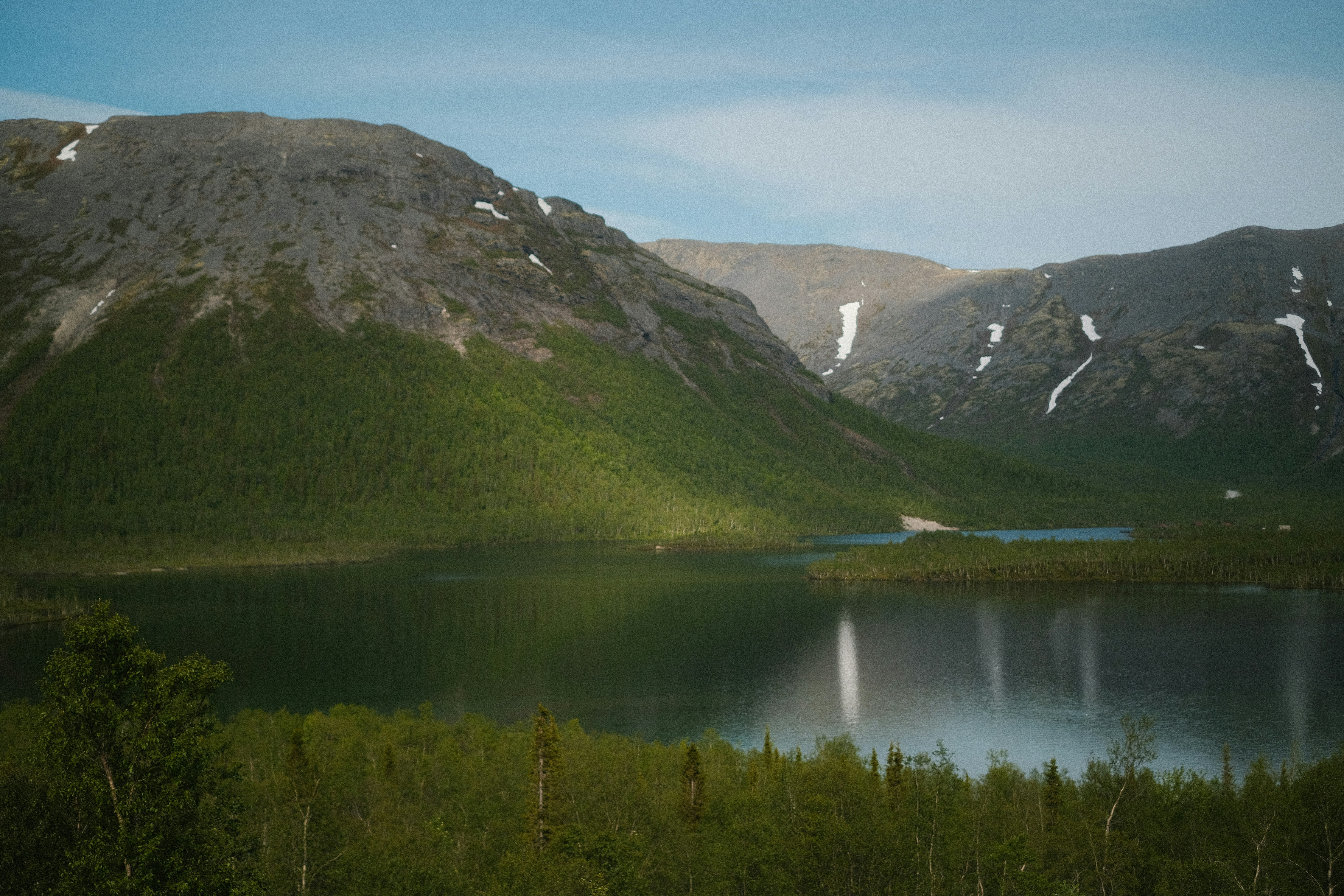 Lush green hills meet tranquil waters, reflecting the rugged mountains under a serene sky. The landscape captures the essence of untouched nature.
