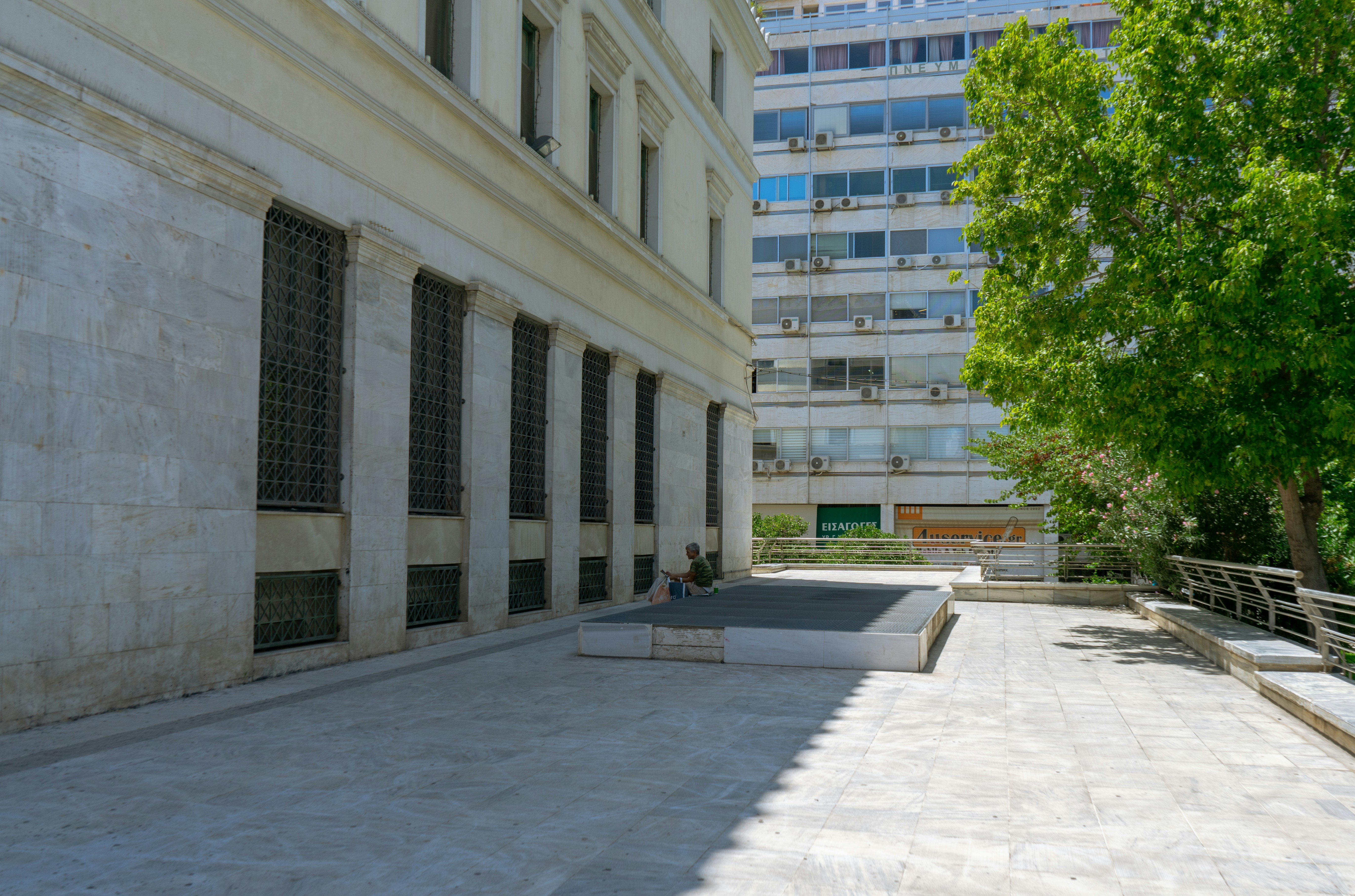 A peaceful urban setting featuring a marble plaza beside a historic building, with a lone figure sitting on a raised platform under the shade of a tree.