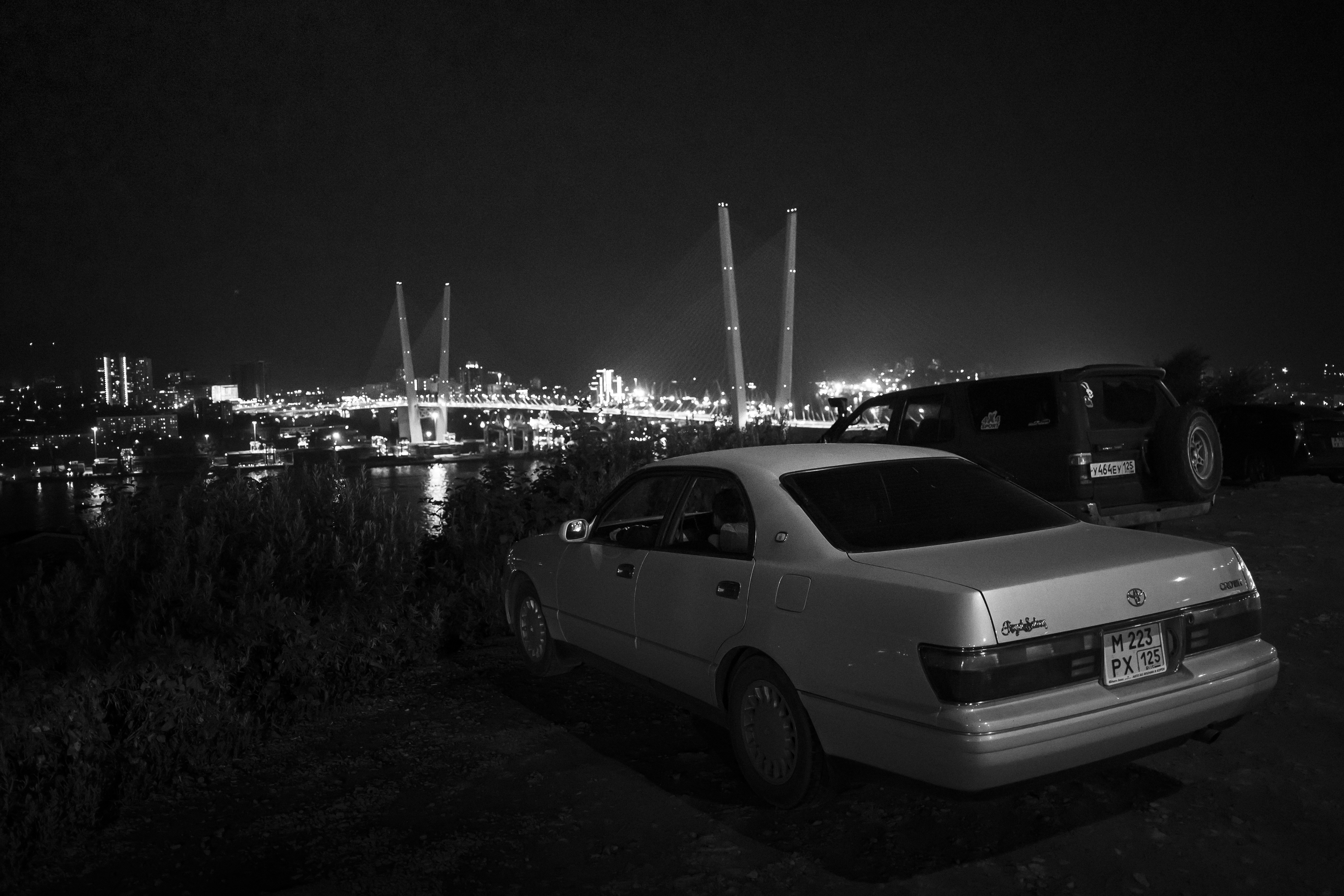 A Japanese vehicle parked on a viewing platform with a view of a suspension bridge in Vladivostok. | A car parks overlooking a city at night.