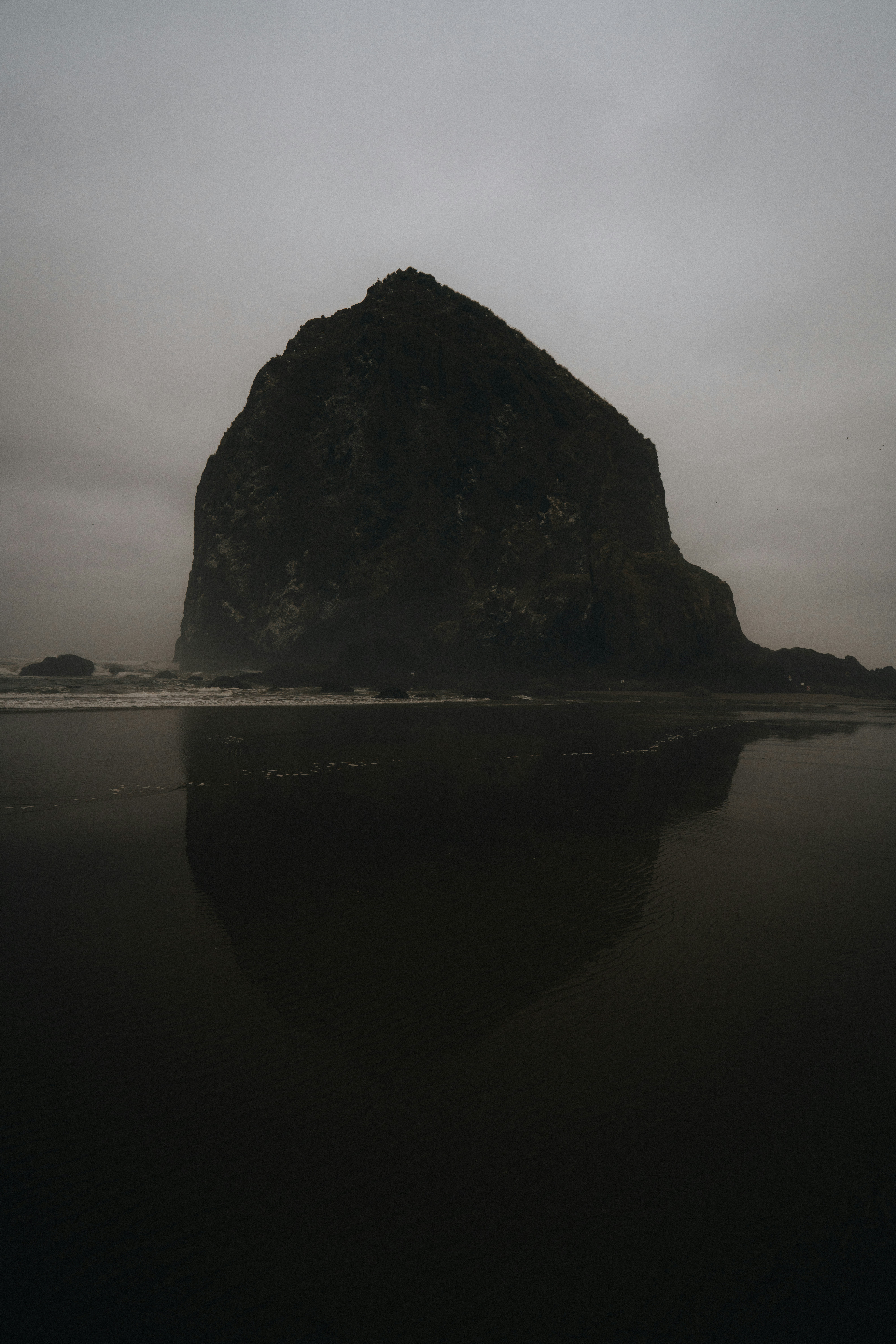 Haystack Rock, Cannon Beach | A dark, moody rock reflected on wet sand.
