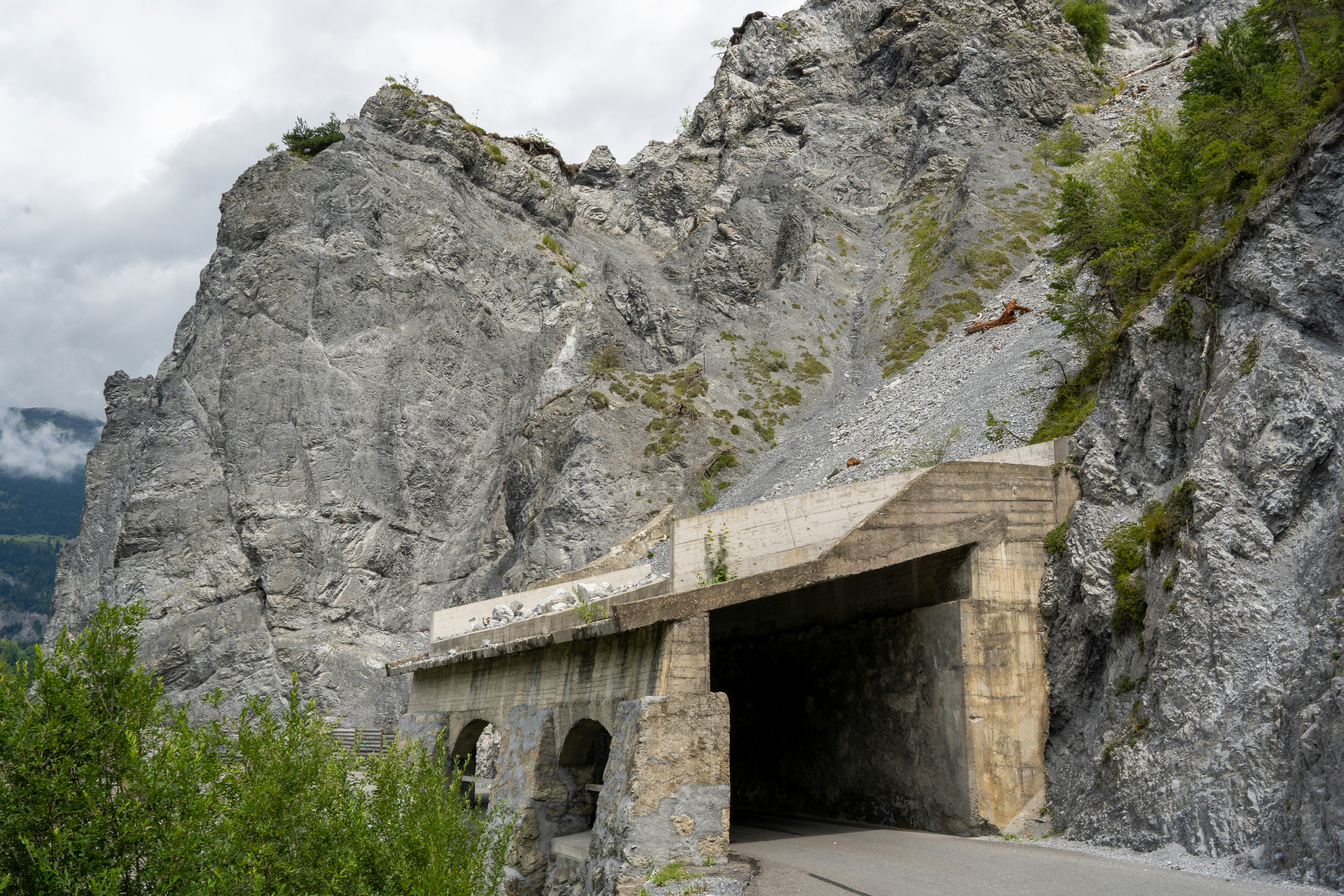 A rugged mountain pass in Graubünden, Switzerland, features a tunnel carved into steep grey rock, partially protected by a weathered concrete overhang. Surrounded by jagged cliffs and sparse alpine vegetation, this dramatic scene showcases the resilience of alpine infrastructure amid raw natural forces. | Tunnel through a rocky mountain.