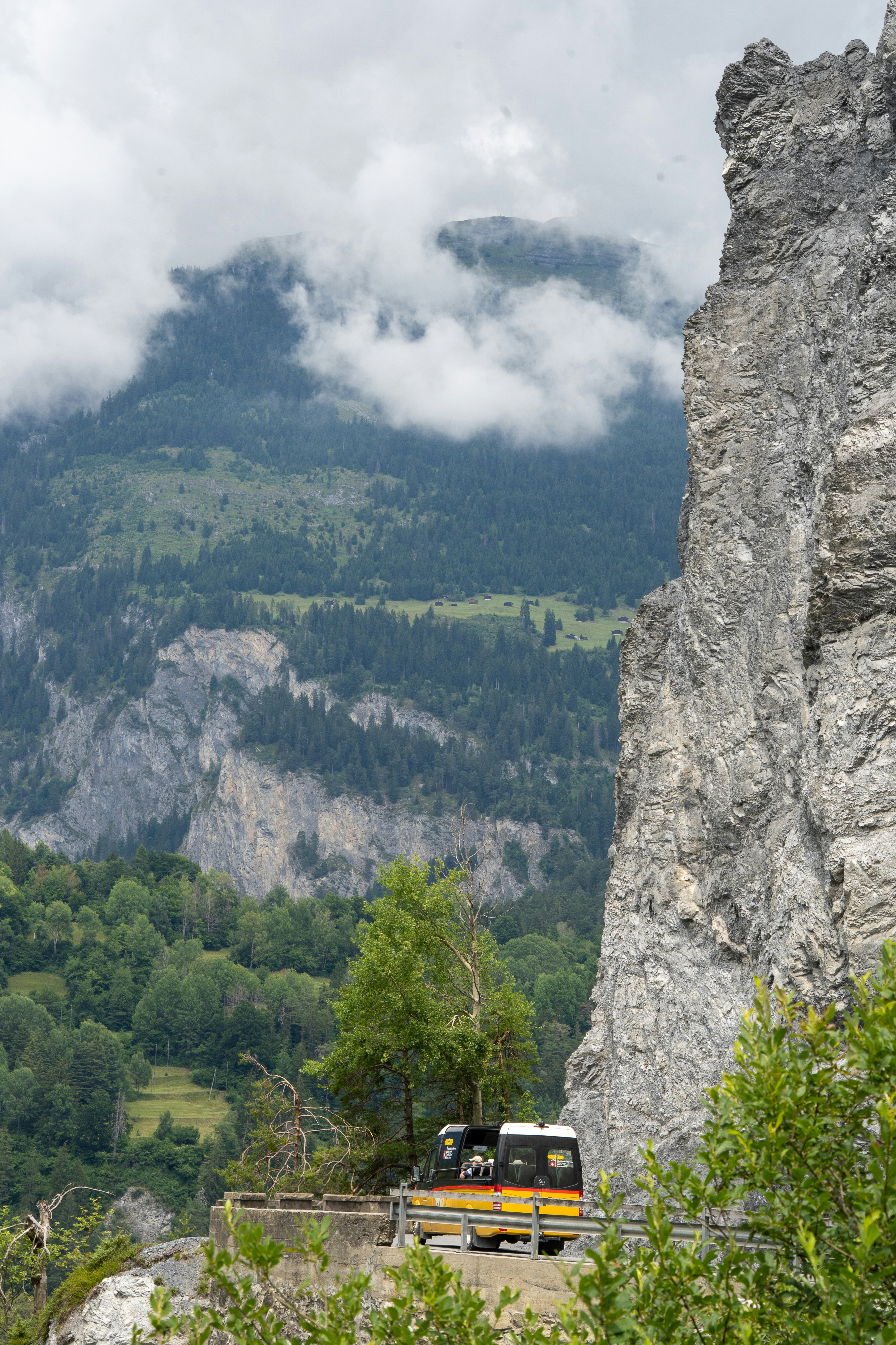 A Swiss PostBus winds along a narrow mountain road near the Vorderrhein in Graubünden, Switzerland, dwarfed by towering cliffs and lush alpine forests. Mist and clouds cling to the peaks, creating a dramatic contrast between rugged rock and vibrant greenery. | A bus drives along a mountain road.