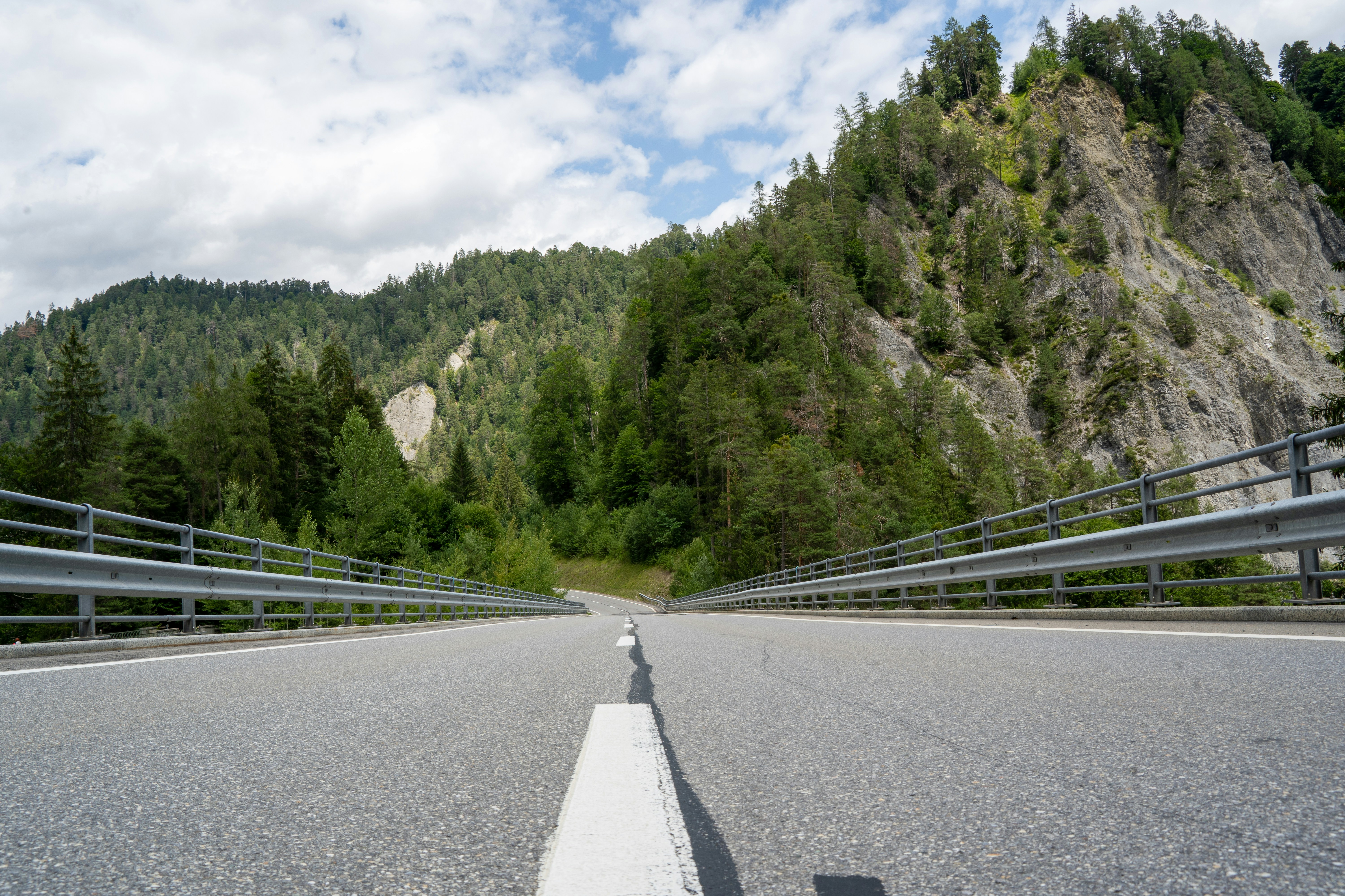 A low-angle view of a mountain road in Graubünden, Switzerland, stretches toward a curve surrounded by towering cliffs and dense evergreen forest. The crisp white line and guardrails frame the scene, guiding the eye into a landscape where alpine nature meets engineered precision. | A road leads towards mountains and trees.