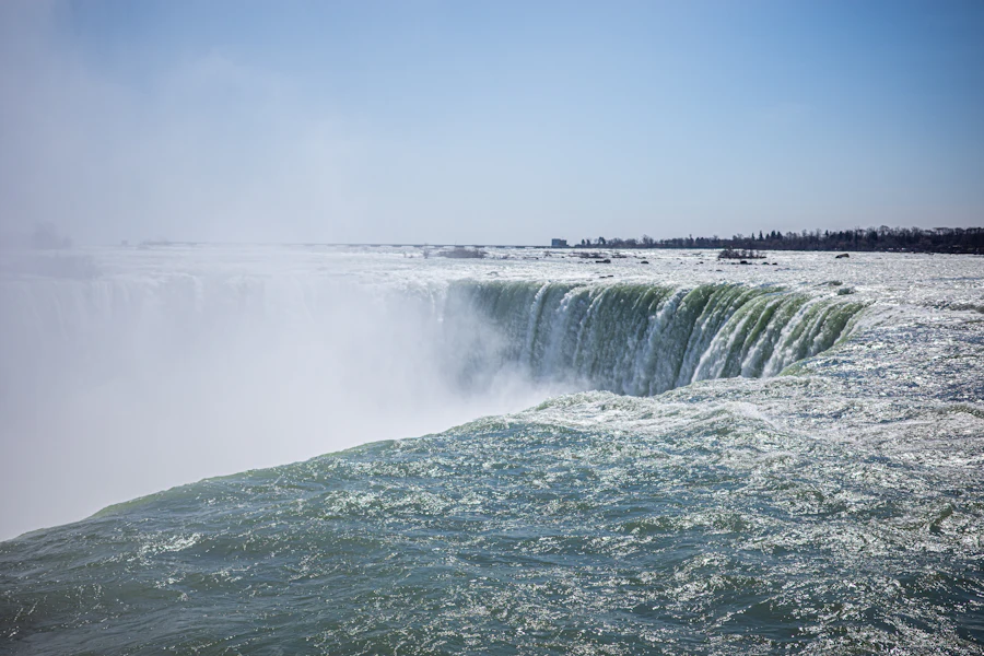 Curving waterfall crest spilling into a mist-filled gorge beneath a pale blue sky.