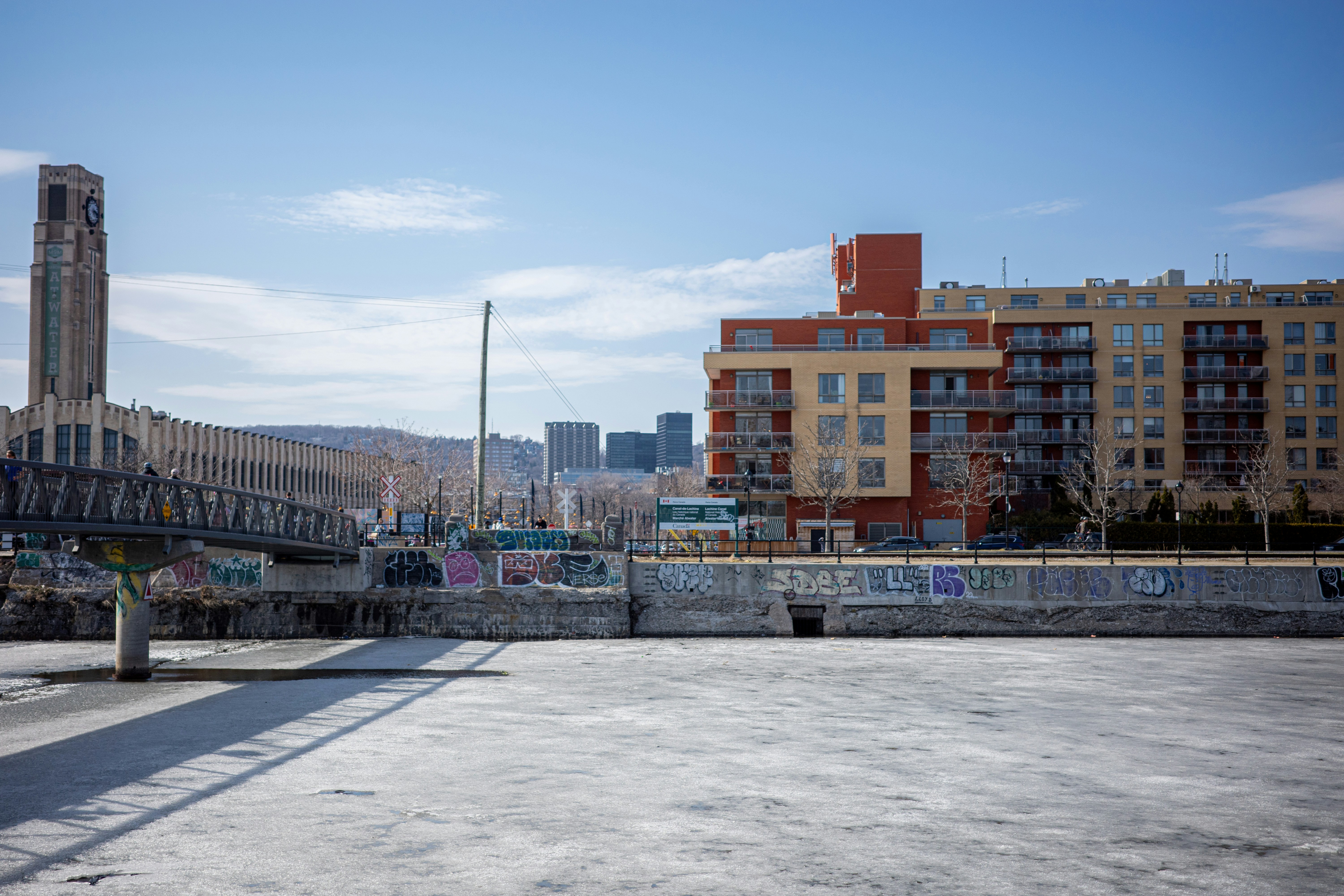 Montreal Winter Bridge Scene with Frozen River and Colorful Apartments. Montreal winter cityscape featuring a bridge crossing the frozen St. Lawrence River with vibrant modern apartment buildings and street art in the foreground. Clear blue winter sky and solid ice surface showcase Montreal's beautiful winter urban landscape. Perfect for Canadian tourism, winter city life, Quebec culture, urban architecture, and cold weather lifestyle content. | A frozen waterfront showcases buildings and a bridge.