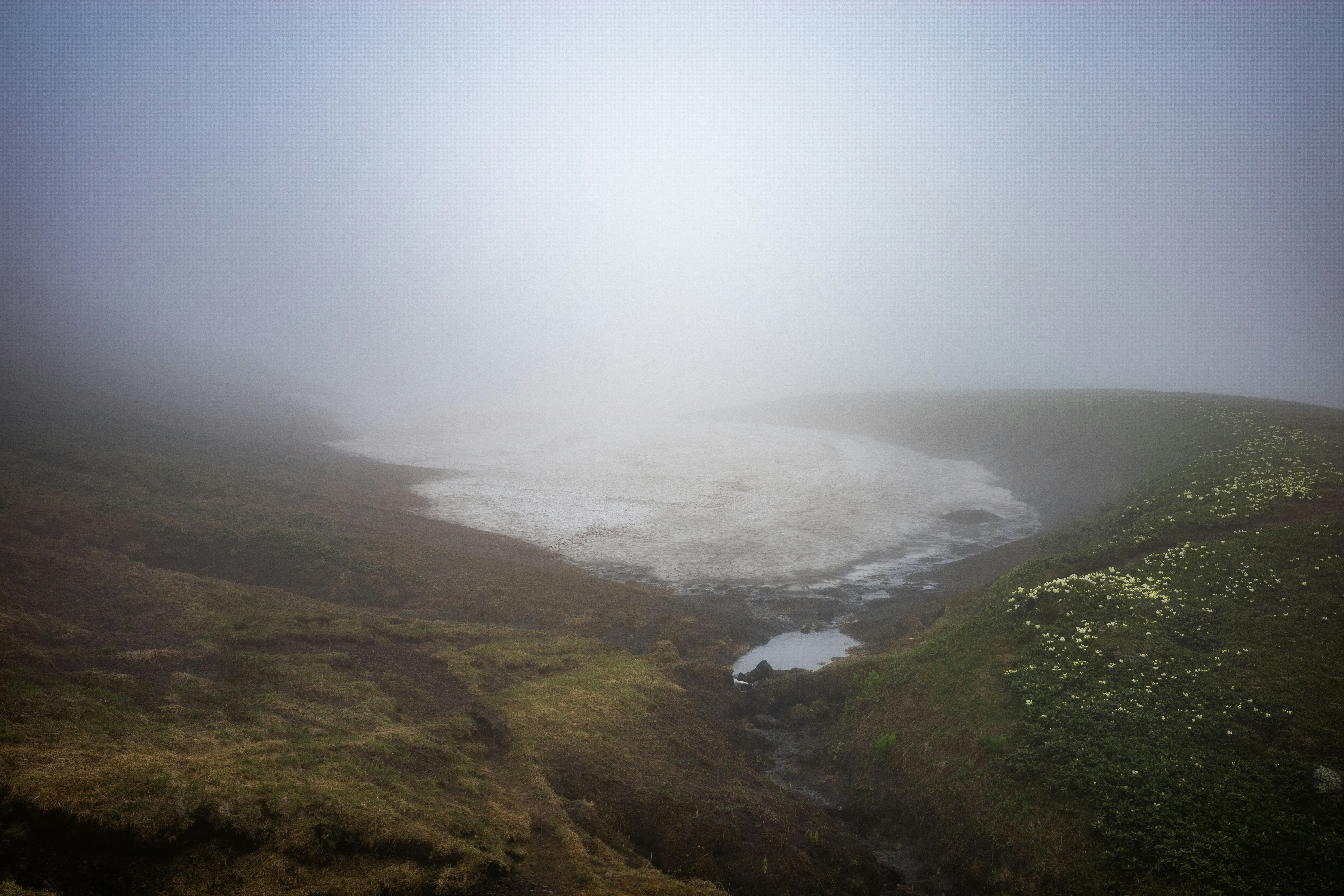 Foggy Day on Mount Paektu Volcanic Landscape in Dense Mist. Mystical foggy landscape on Mount Paektu shrouded in thick mist on the North Korea-China border. The sacred volcanic mountain emerges through dramatic fog in this atmospheric view of the highest peak on the Korean Peninsula. Perfect for volcanic landscape photography, East Asian mountain tourism, atmospheric weather imagery, and sacred mountain themes. | Misty mountain landscape with remnants of snow.