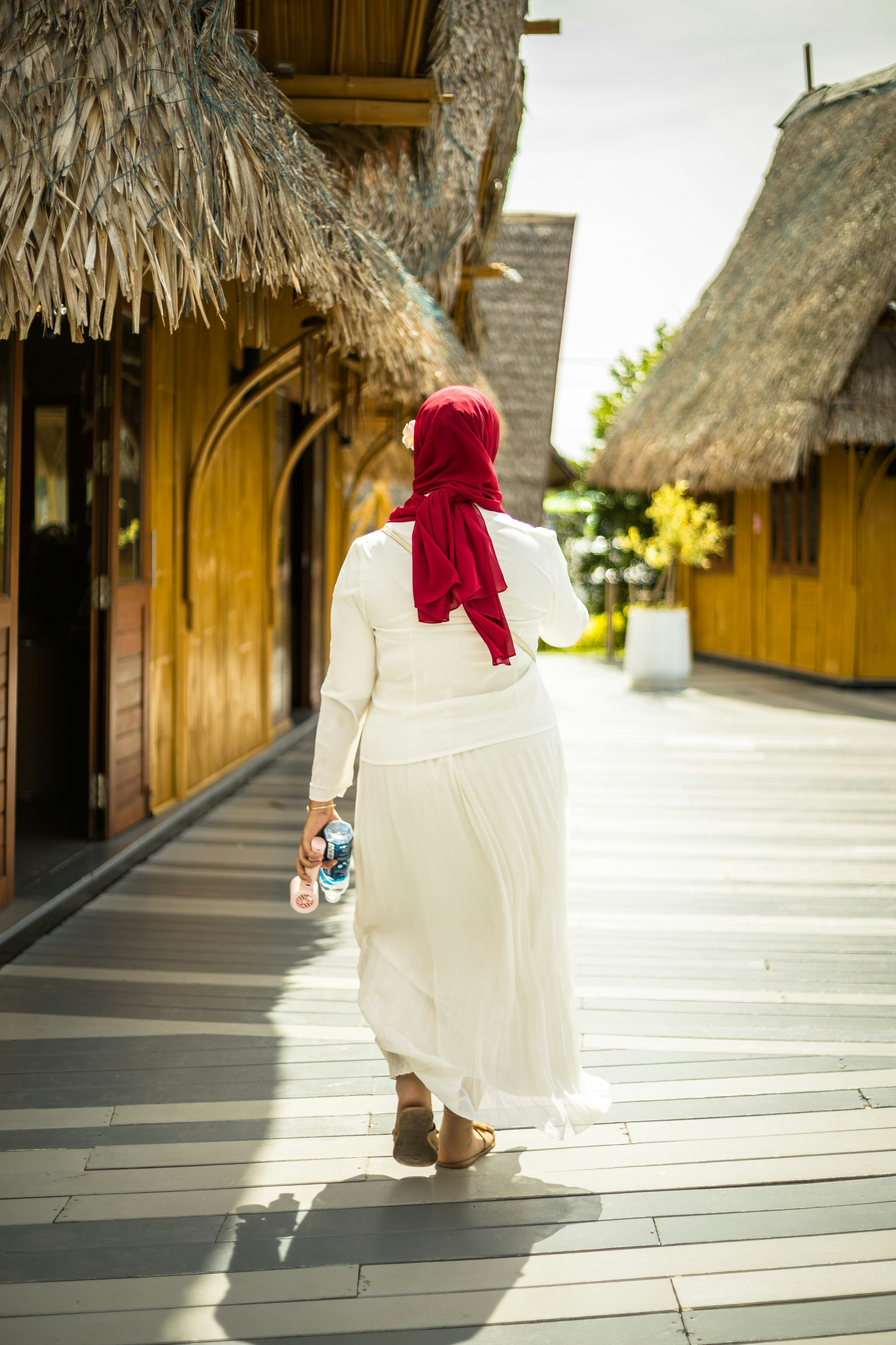 Woman in a flowing white dress and red scarf walking along a wooden pathway lined with thatched-roof structures.