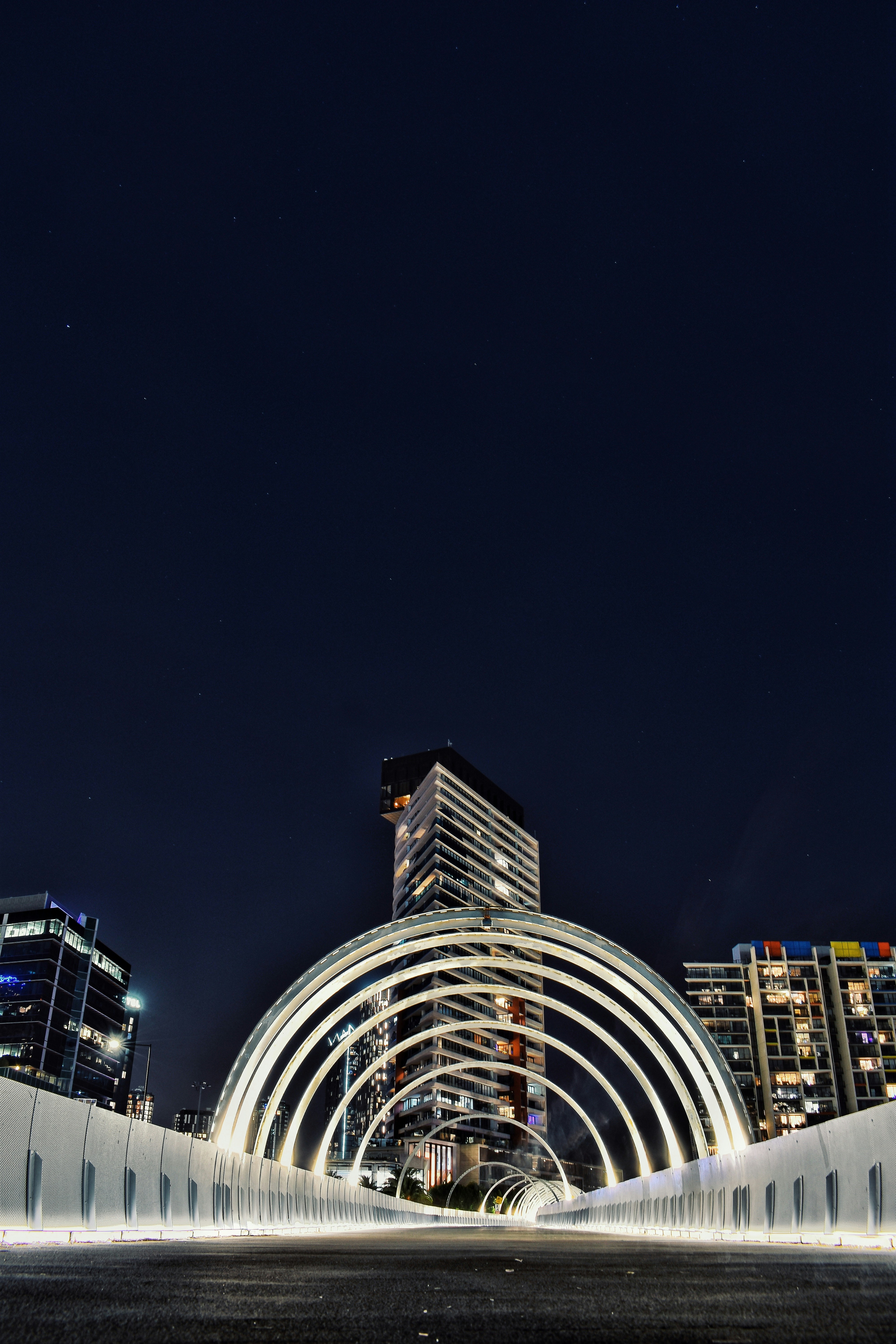 Architectural bridge featuring illuminated arches against a night sky, framed by city buildings. The scene conveys a sense of movement and modernity.