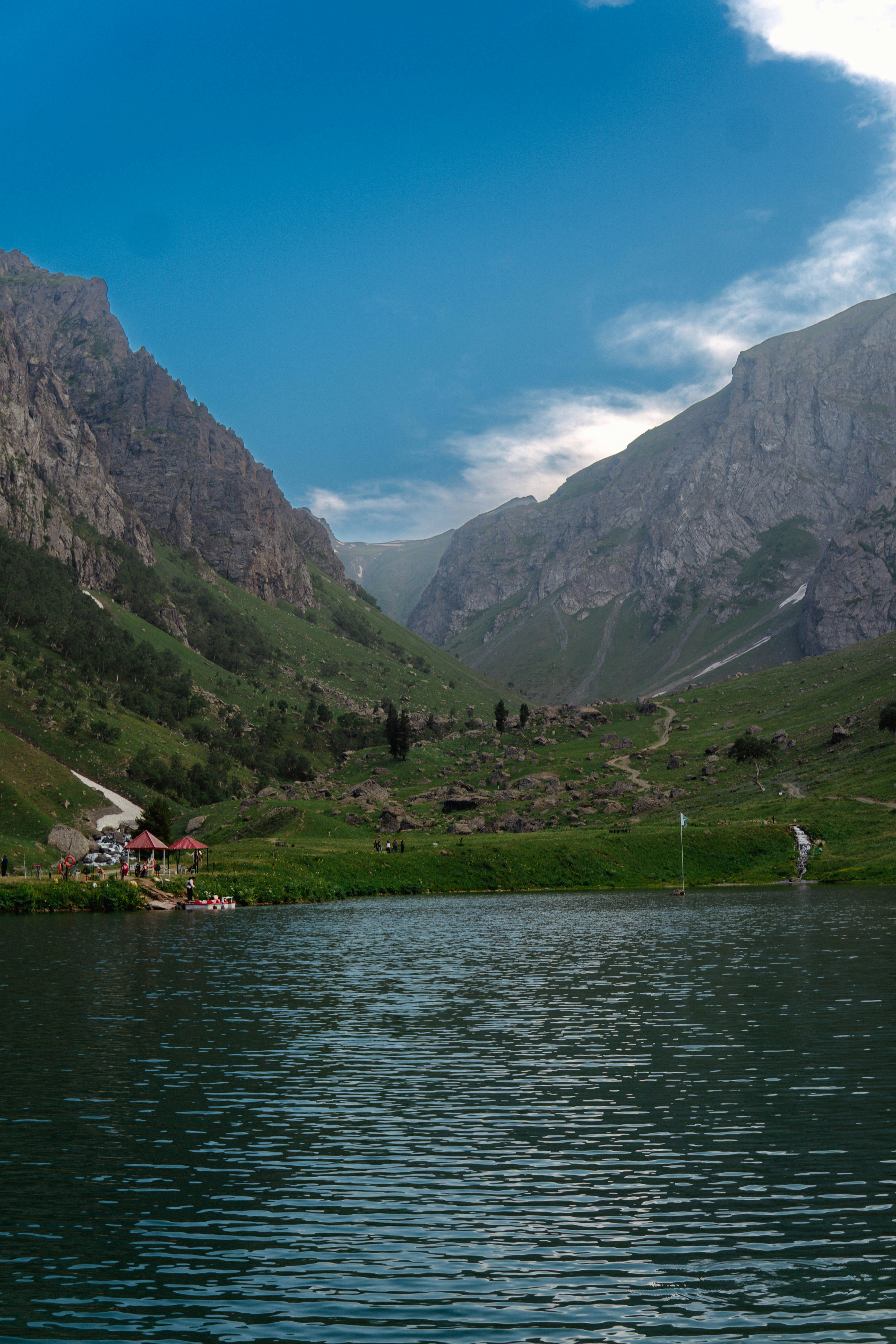 Serene lake nestled between towering mountains, dotted with lush greenery and a quaint building by the shore.