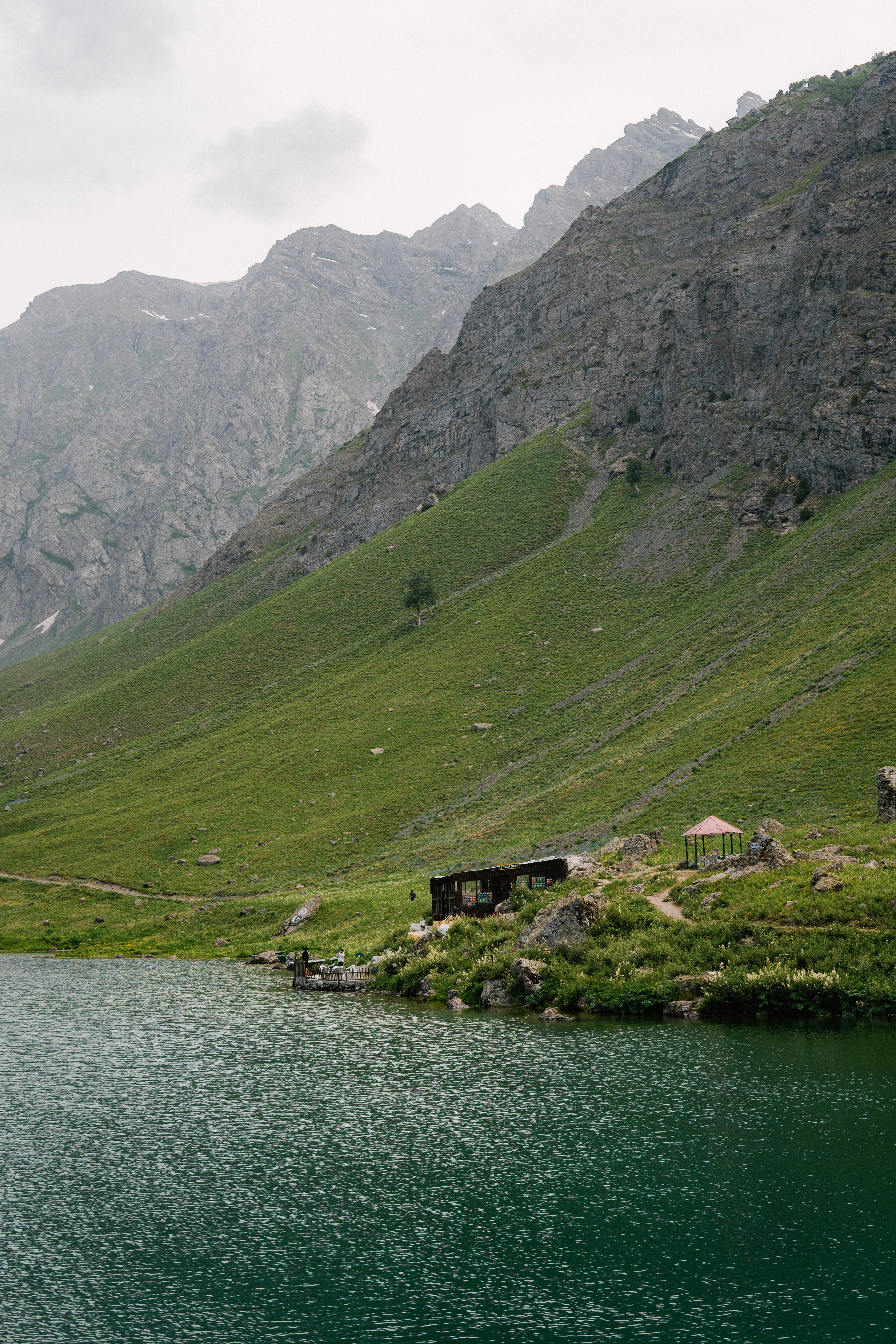 Rustic cabin nestled by a serene lake, surrounded by lush green hills and towering mountains. Ideal for tranquil escapes.