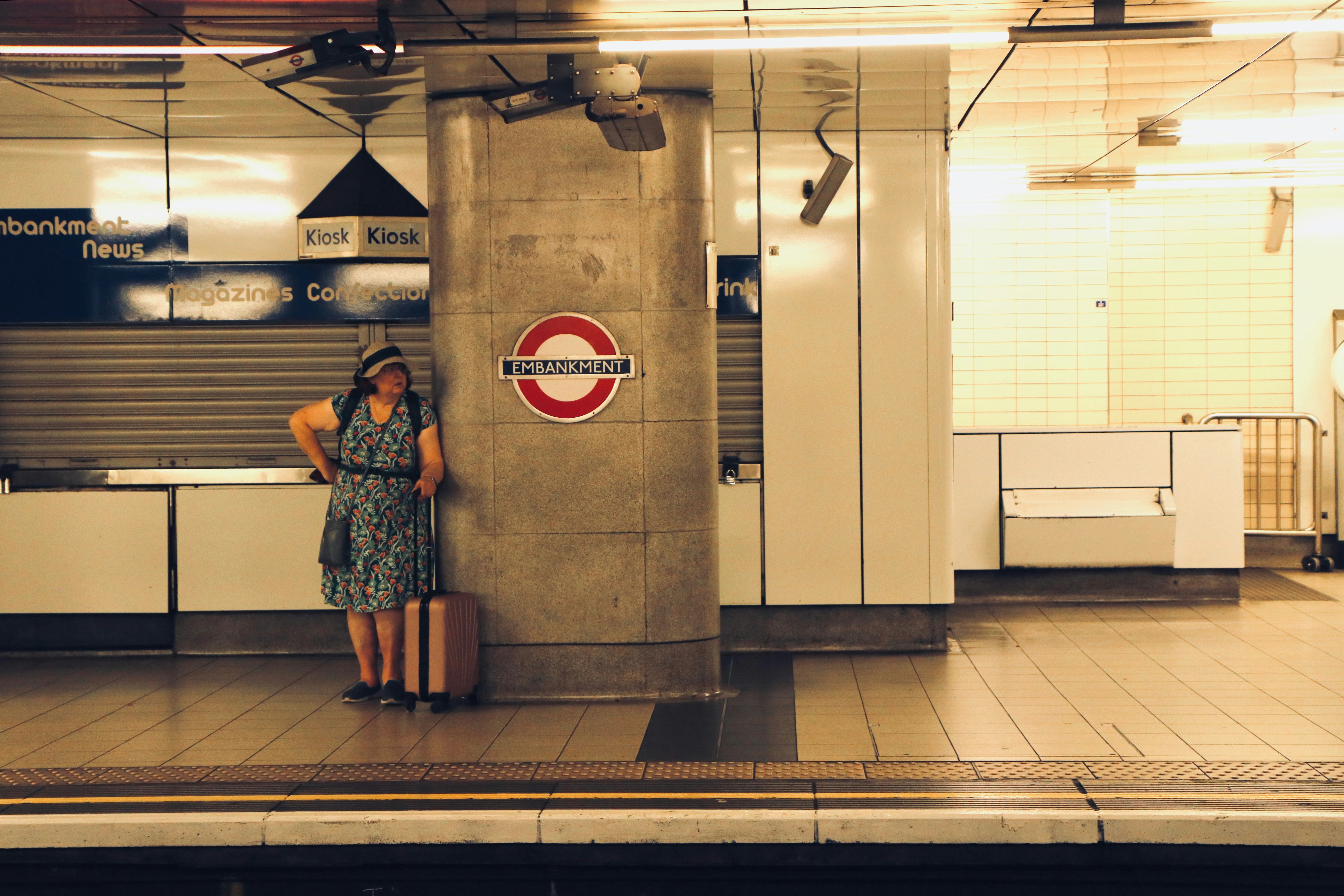 A woman in a floral dress stands beside a luggage at a subway station, leaning against a concrete pillar adorned with a circular transit logo.