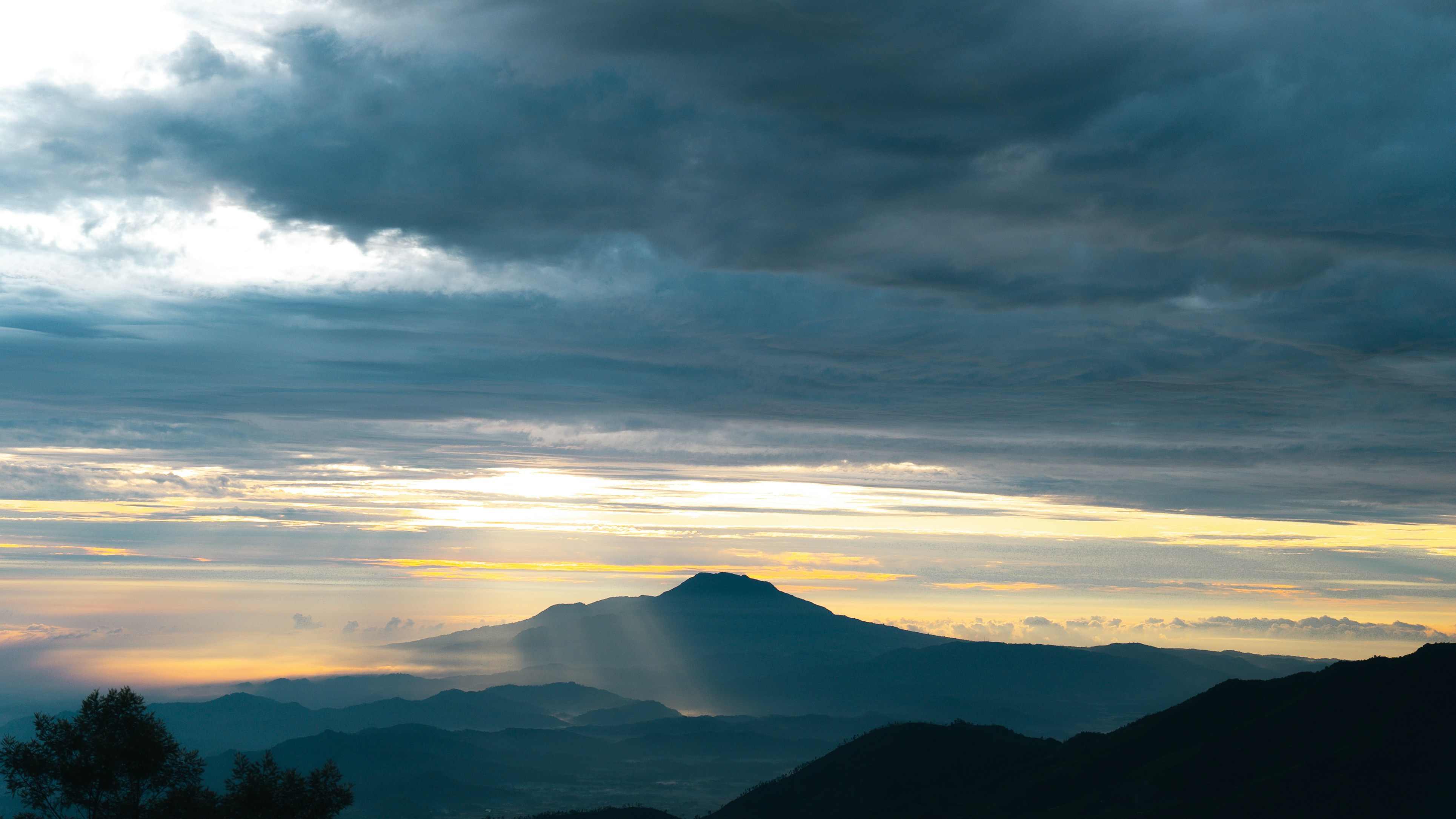 Mountains silhouetted against a dramatic sky as the sun sets, casting rays through the clouds. The serene landscape evokes a sense of tranquility.