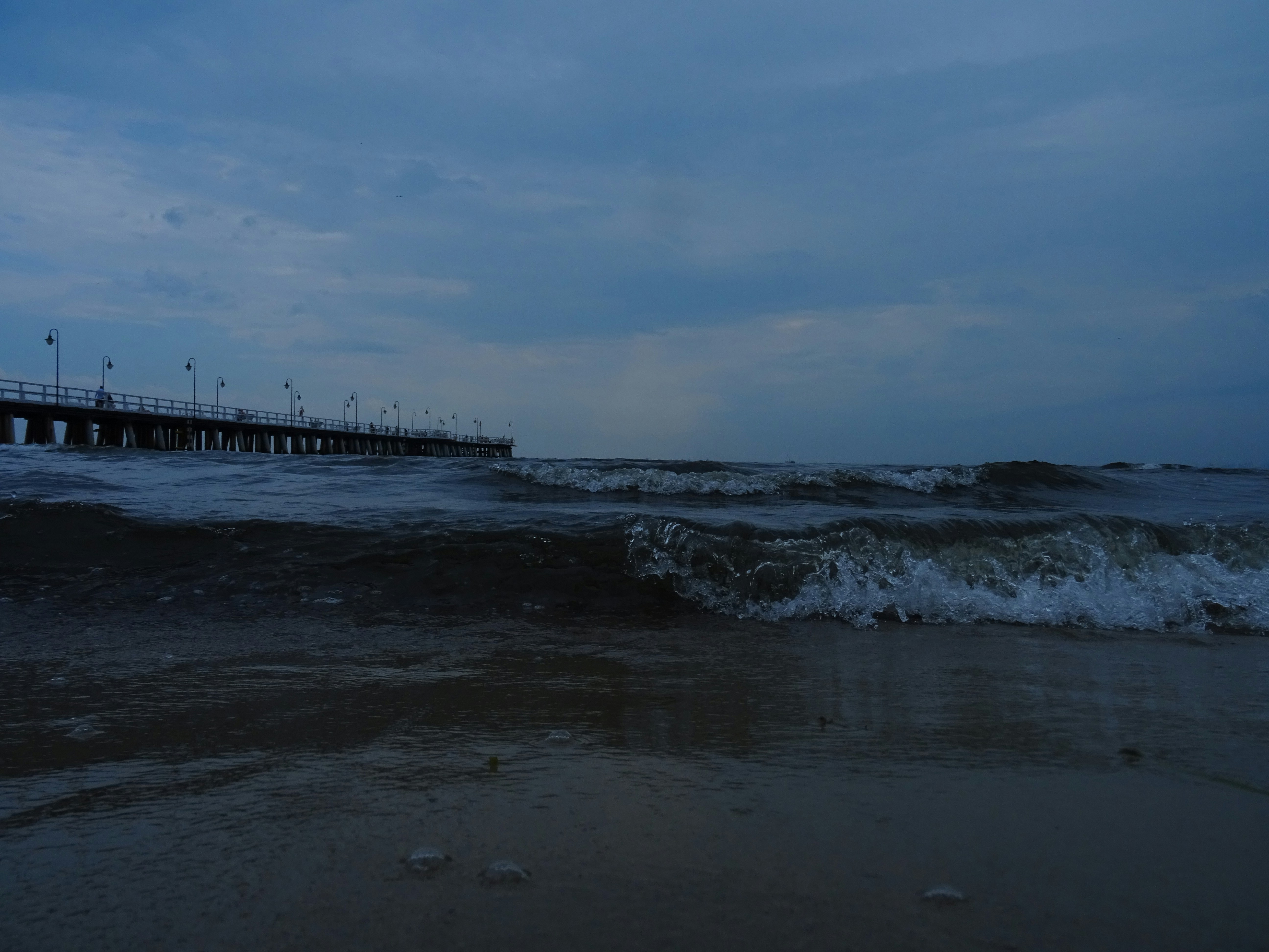 Waves crash near a pier under a cloudy sky.
