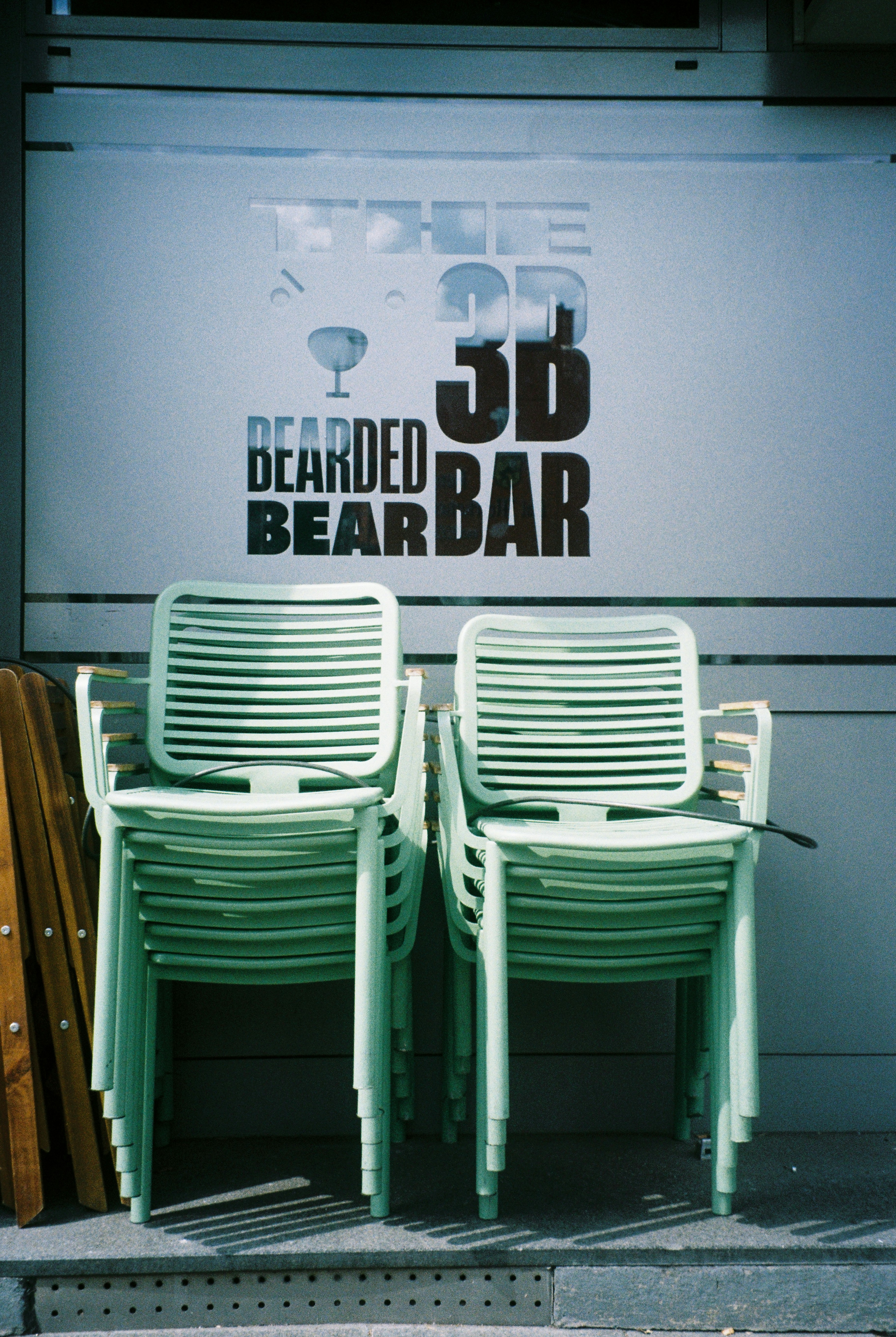 Stacked mint green chairs in front of a playful bar sign featuring a bear motif. The scene captures a casual outdoor setting.