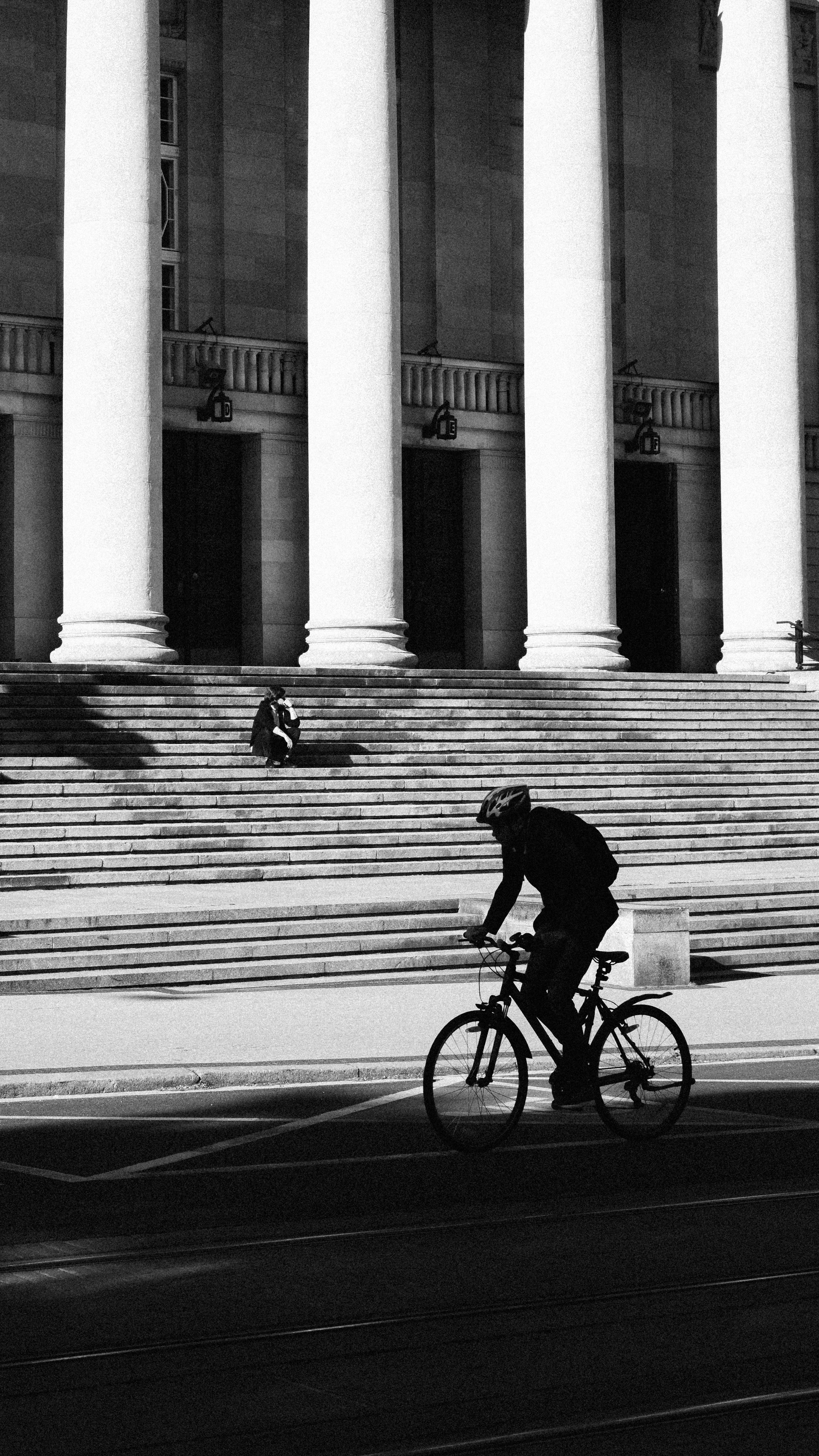 A cyclist speeds past grand columns while a figure rests on the steps, embodying the contrast between movement and stillness in an urban environment.