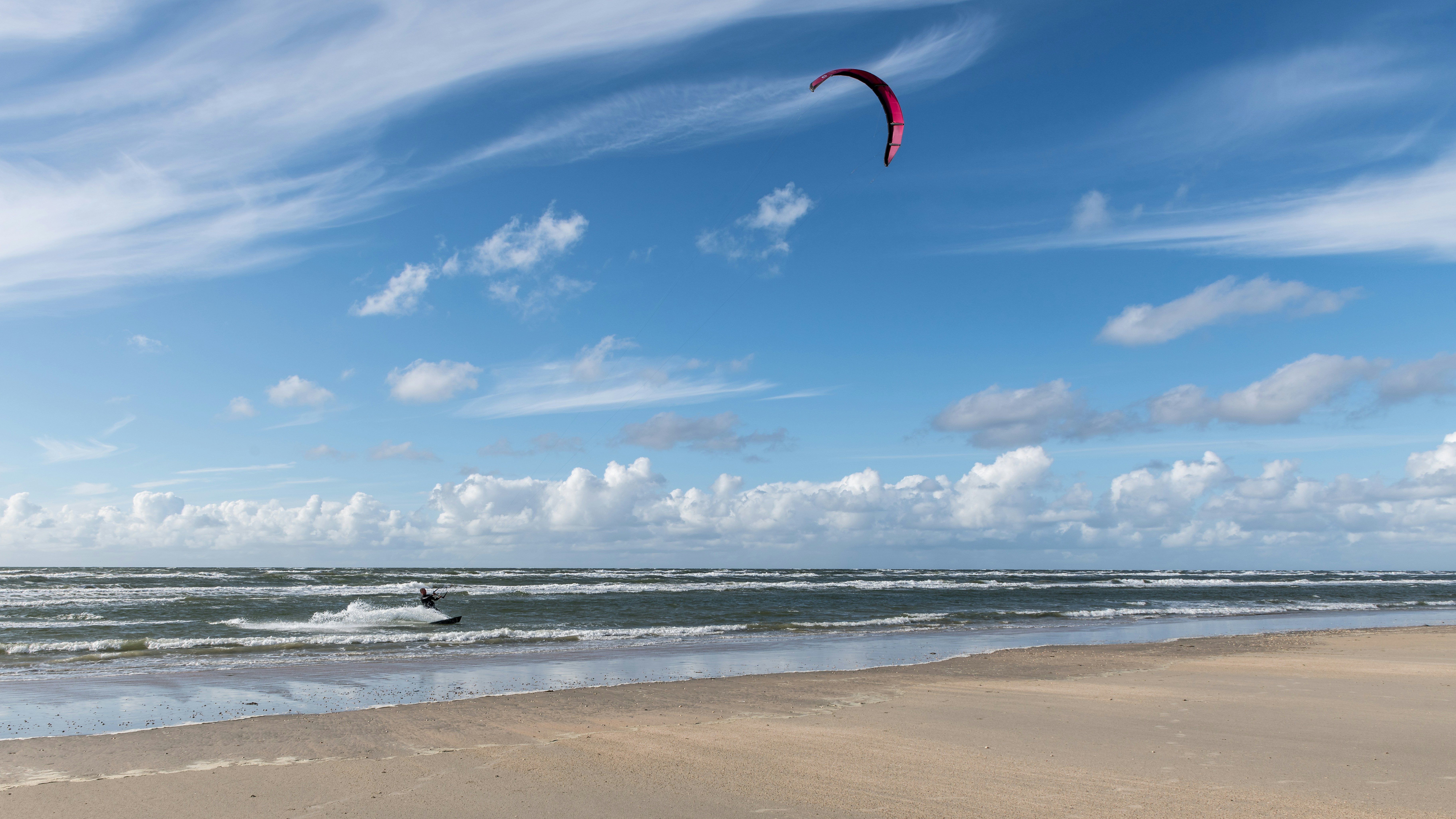 A kite surfer gracefully navigates the waves under a vibrant sky filled with wispy clouds and a colorful kite soaring above. 