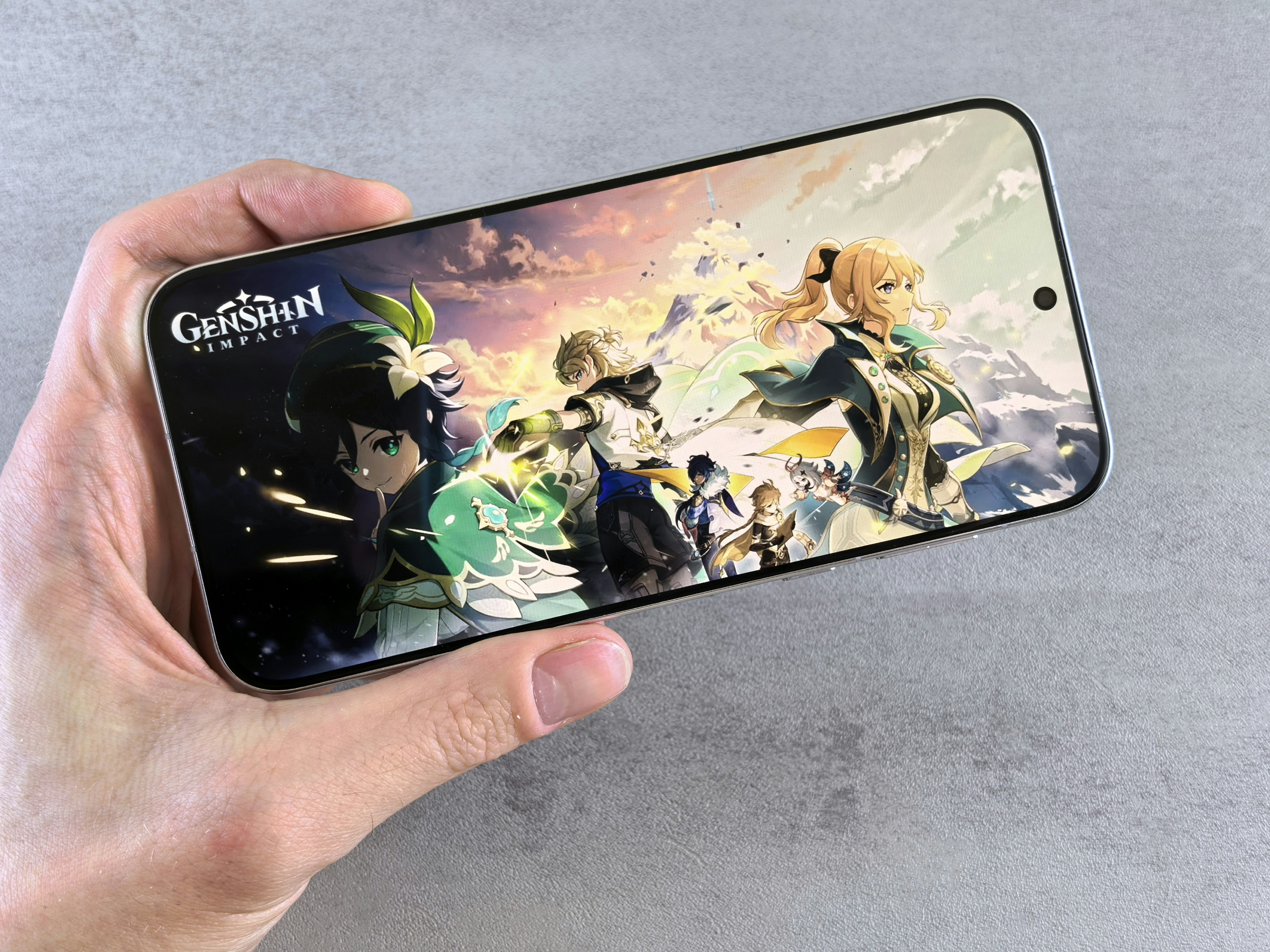 A hand holds a smartphone displaying vibrant artwork from the game Genshin Impact, showcasing characters against a dramatic sky.
