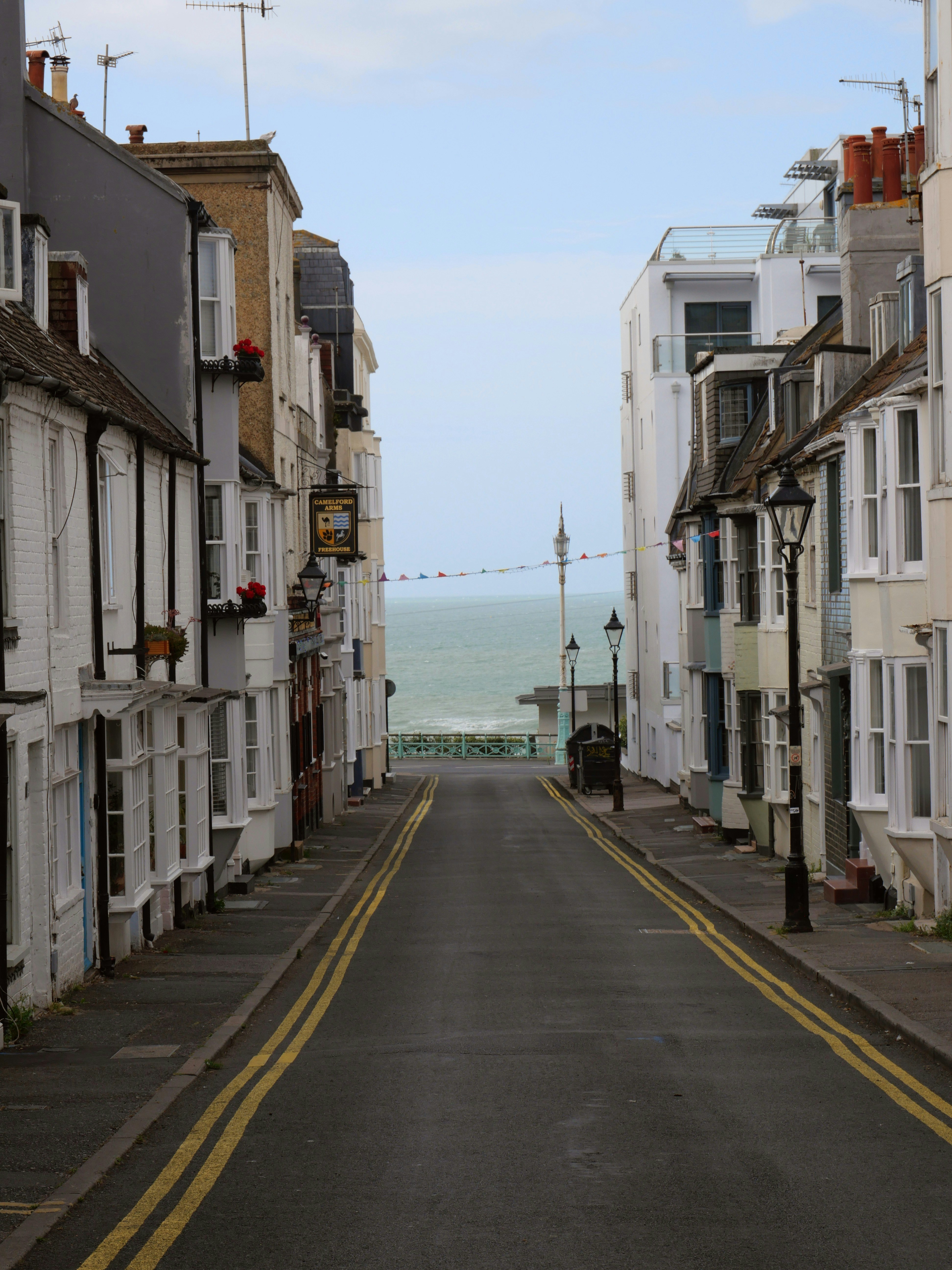 Eine enge Gasse in Brighton, Großbritannien, mit charakteristischen Gebäuden, die zum Meer hinabführen, welches am Horizont sichtbar ist. Die gepflasterte Straße und die doppelten gelben Linien vermitteln einen urbanen Cha | A street leads to the sea.