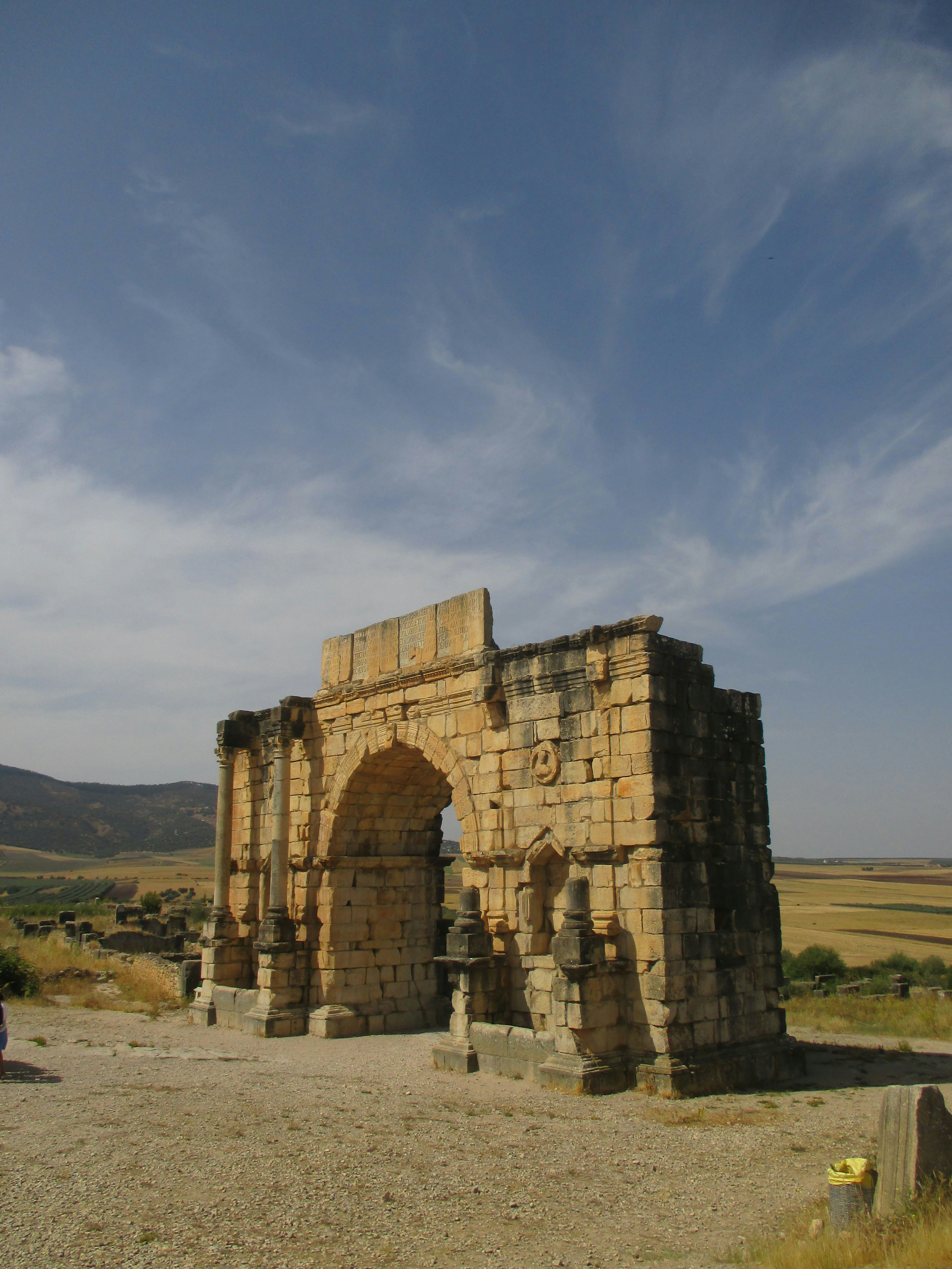 Volubilis Arc de Triomphe Maroc | Ancient ruins stand under a blue sky.