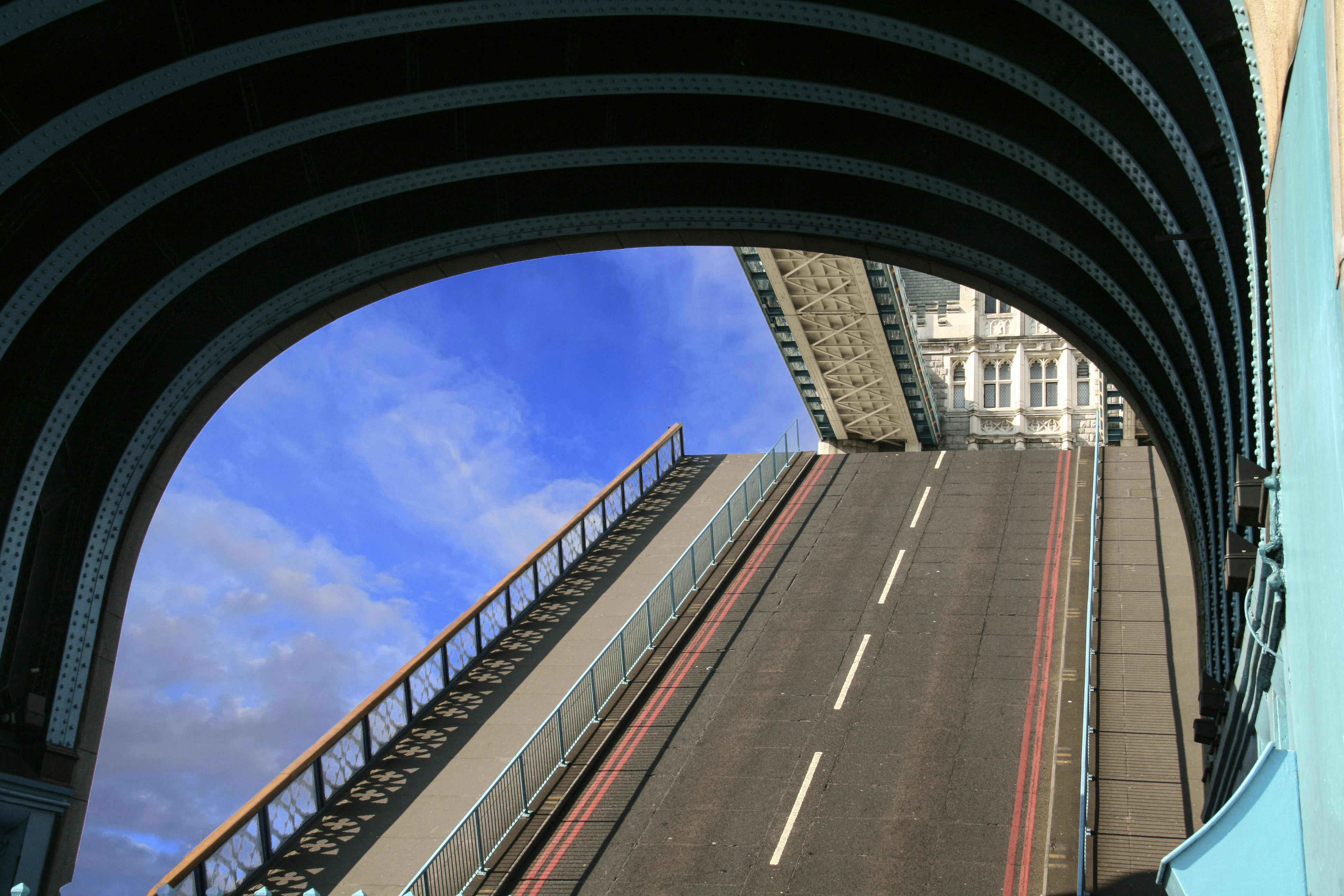 This striking photo captures Tower Bridge in London with its bascules fully raised to allow the passage of a vessel on the River Thames. Framed from beneath the bridge, the image emphasizes the engineering marvel and symmetry of the structure, highlighting the iconic red and white road markings against the vivid blue sky. | Tower bridge's roadway is raised, revealing sky.