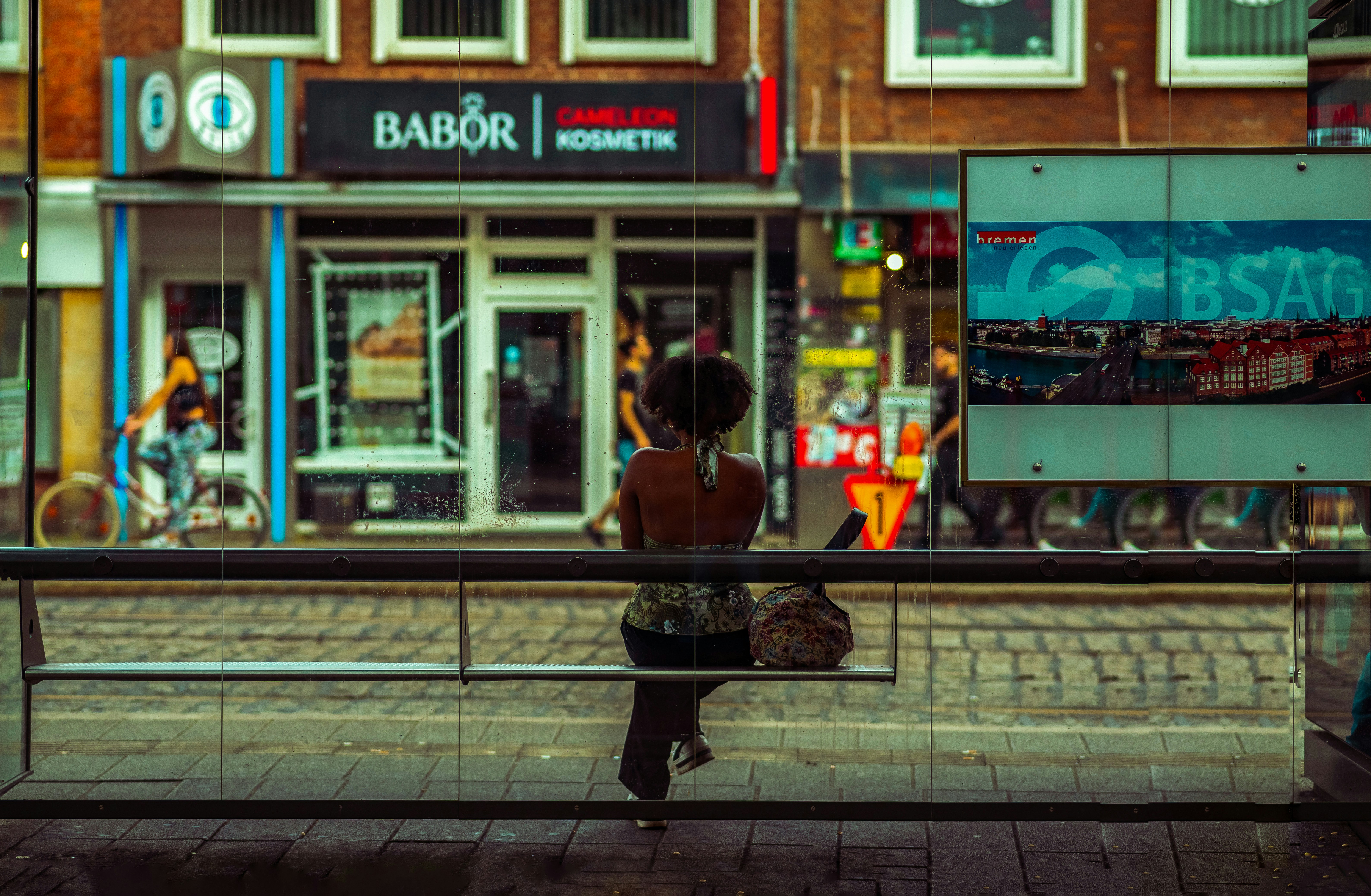 A solitary figure sits at a bus stop, framed by glass, watching the bustling street scene outside. The contrasting reflections create a layered visual narrative.