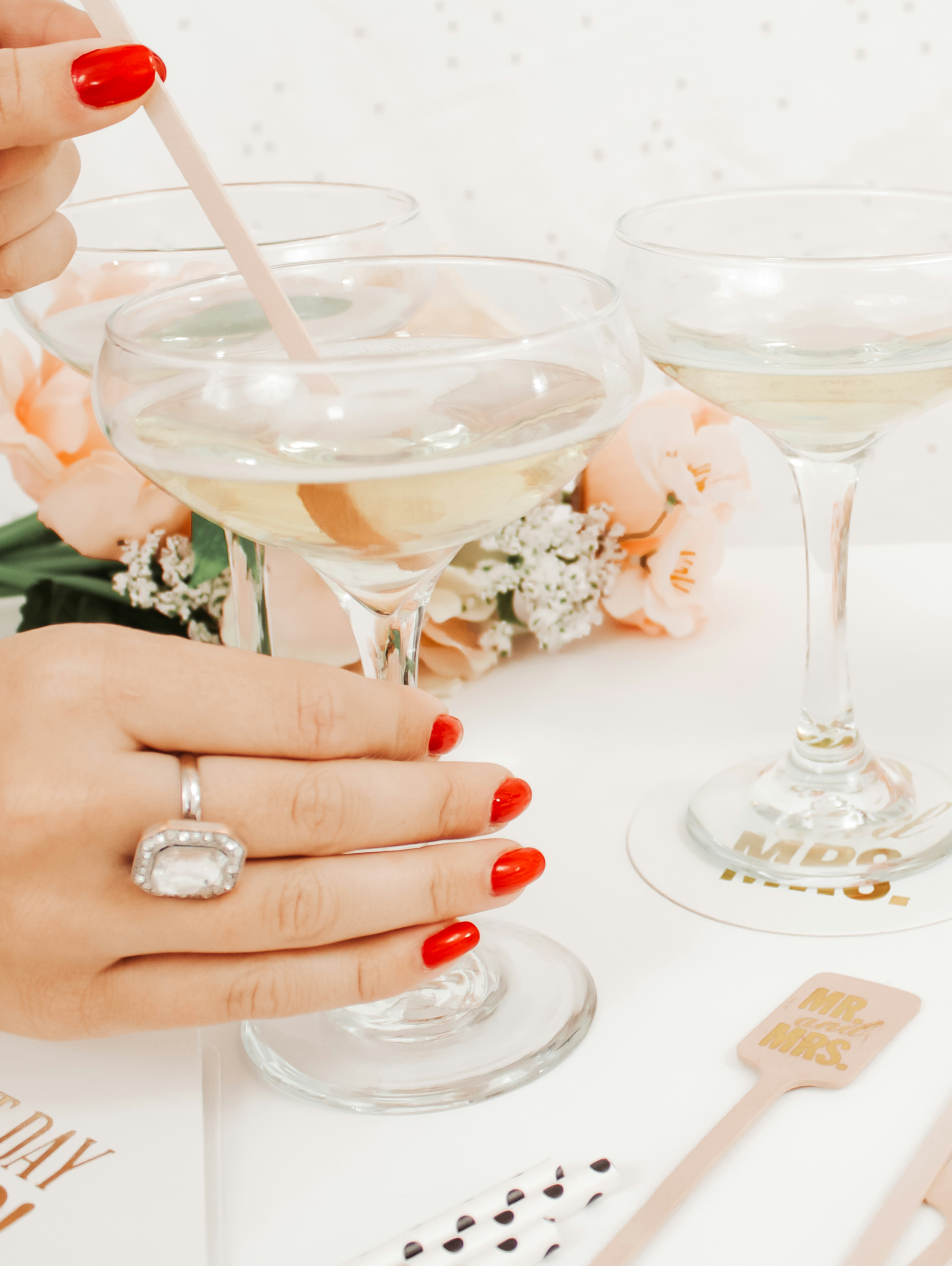 A hand with striking red nails stirs a cocktail in a glass, surrounded by elegant floral arrangements and decorative stirrers, celebrating a special occasion.