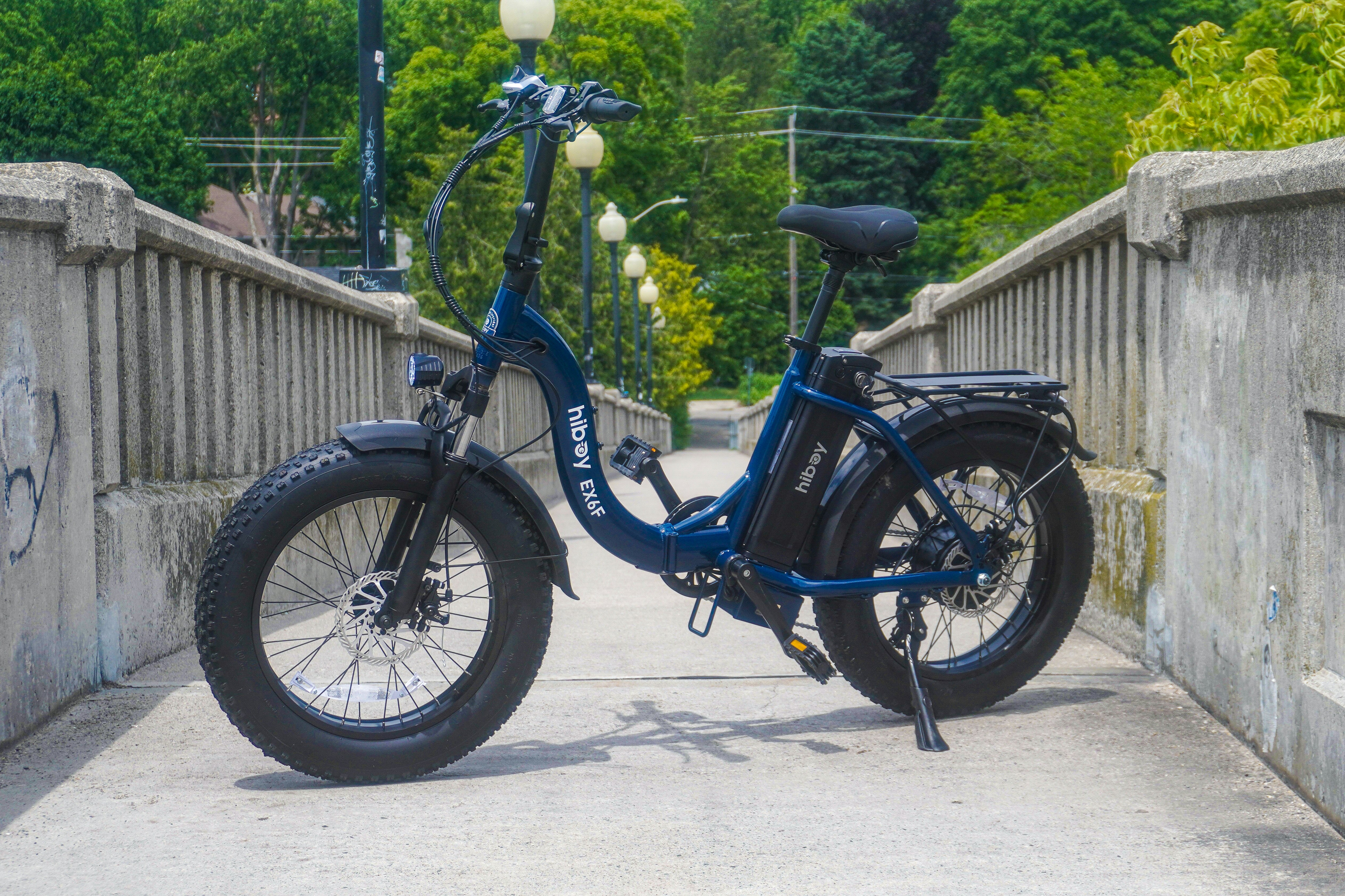 Electric fat bike parked on a bridge, surrounded by lush greenery and urban elements. The bike's robust design emphasizes adventure and mobility.