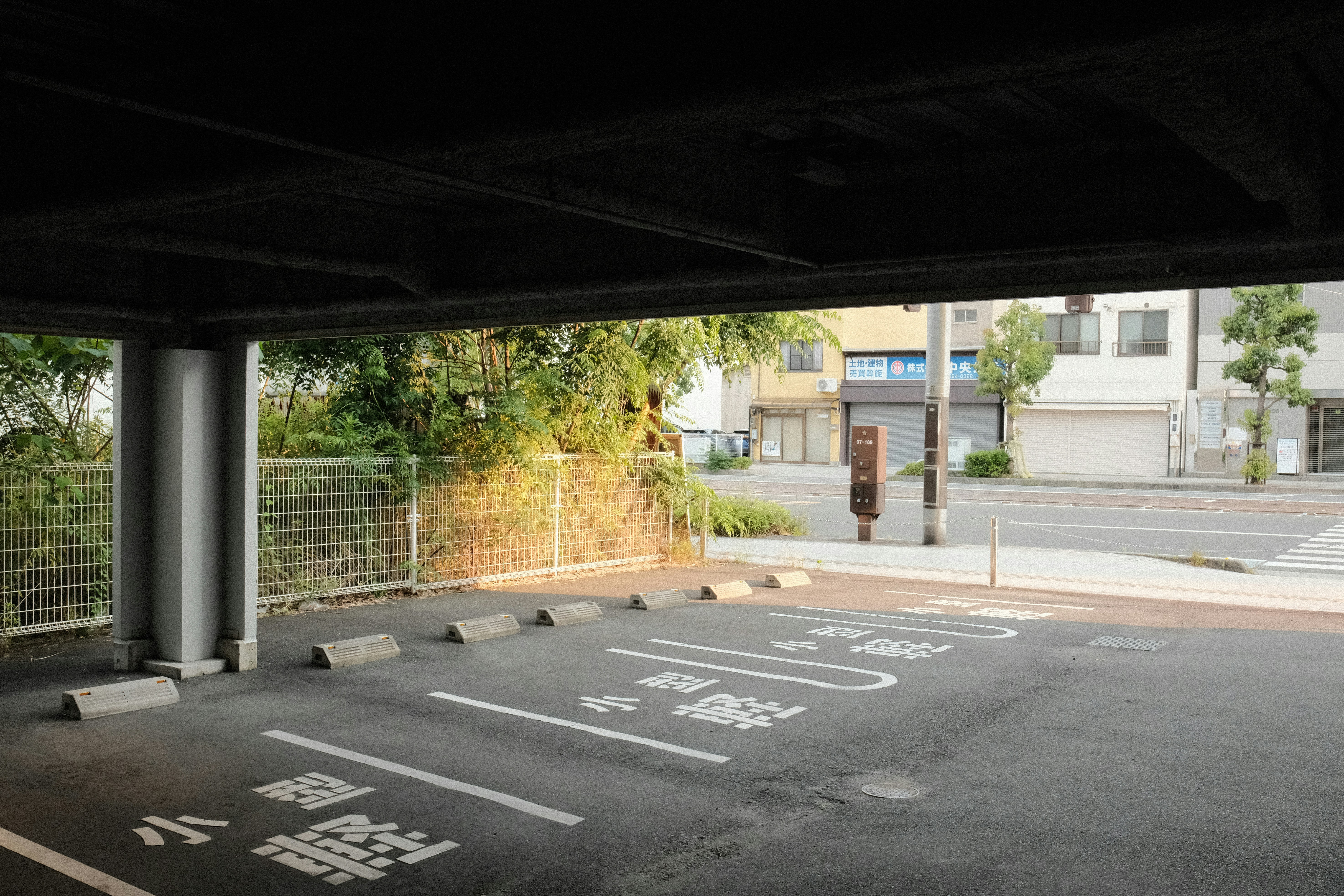 Empty parking lot under a structure, with greenery peeking through the fencing and buildings in the background. The scene captures urban tranquility.