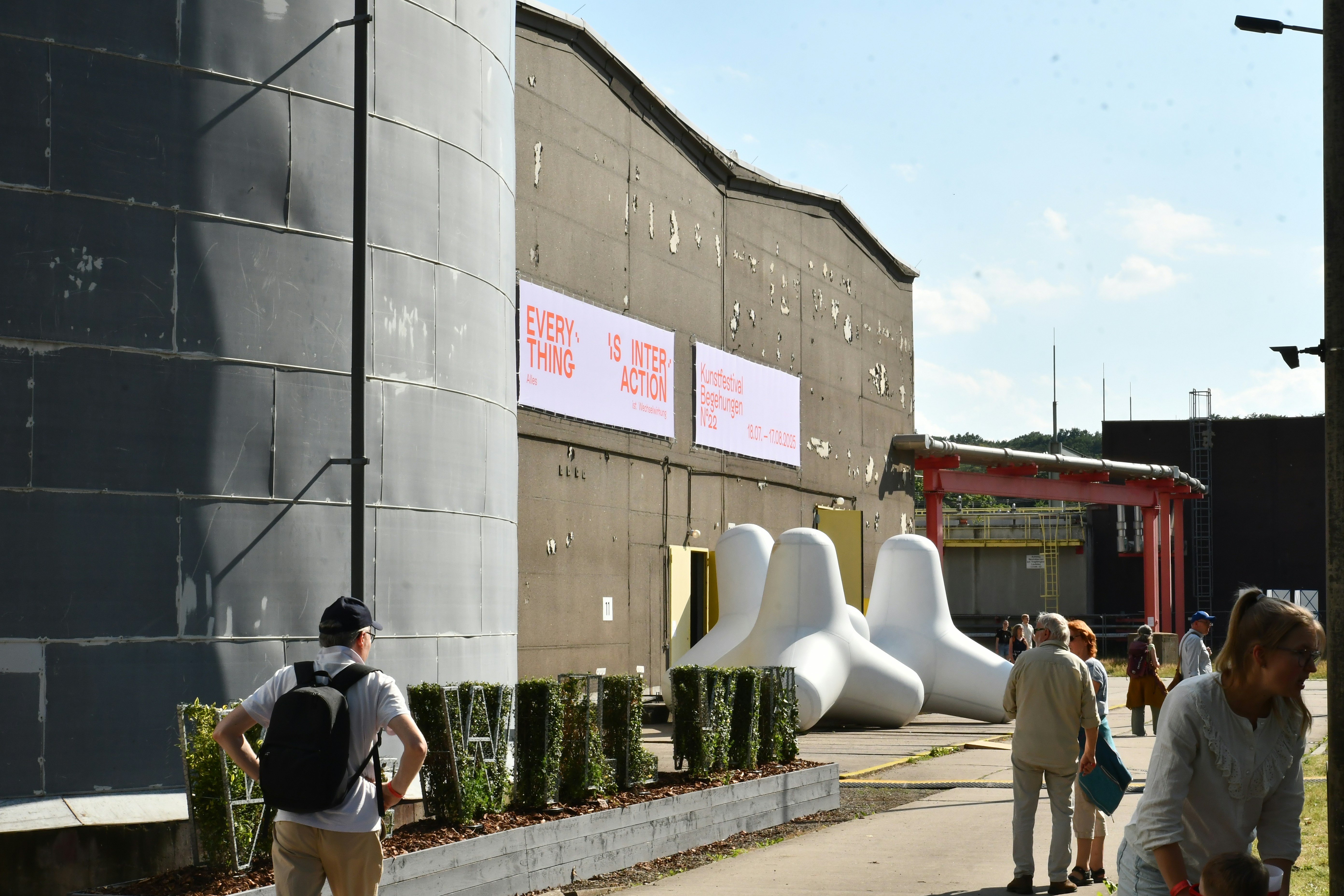 People walk by industrial buildings on a sunny day.