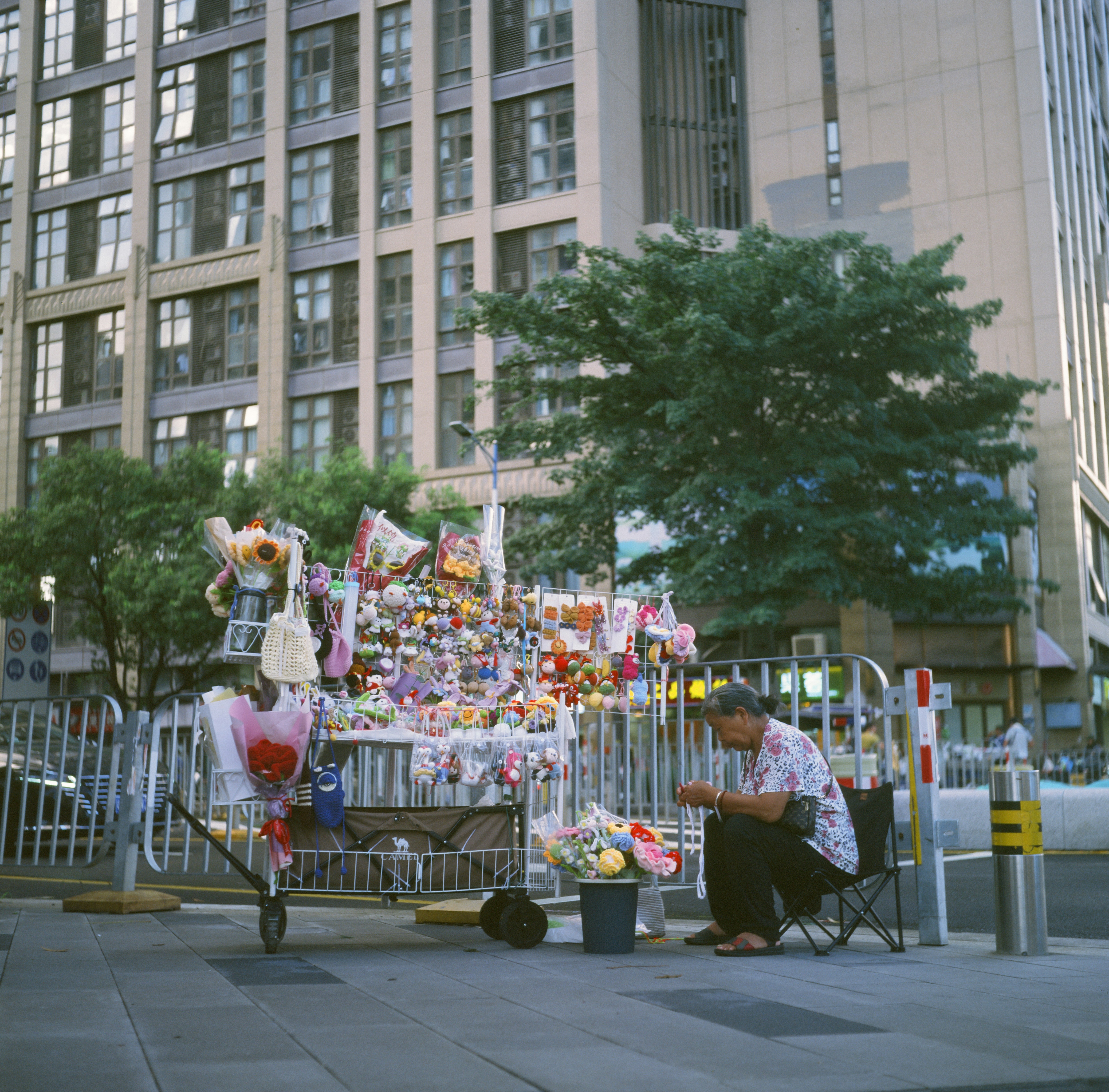 This is another small street stall selling trinkets and accessories. When no customers are around, the vendor passes the time by knitting little crafts. | A street vendor selling wares sits in the city.