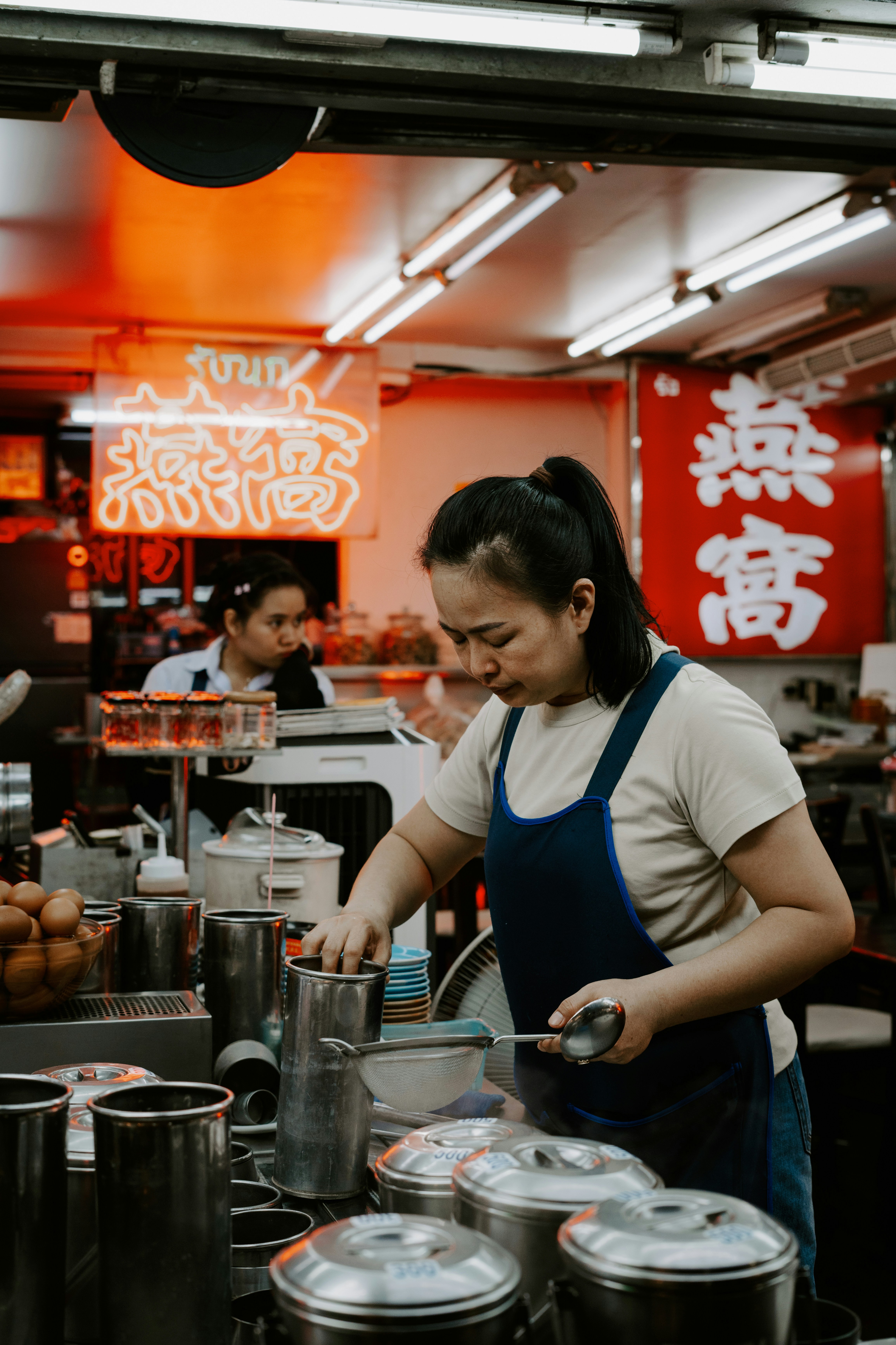 A vendor meticulously preparing food in a bustling market, surrounded by vibrant neon signs and various cooking utensils. The atmosphere reflects the lively energy of street food culture.