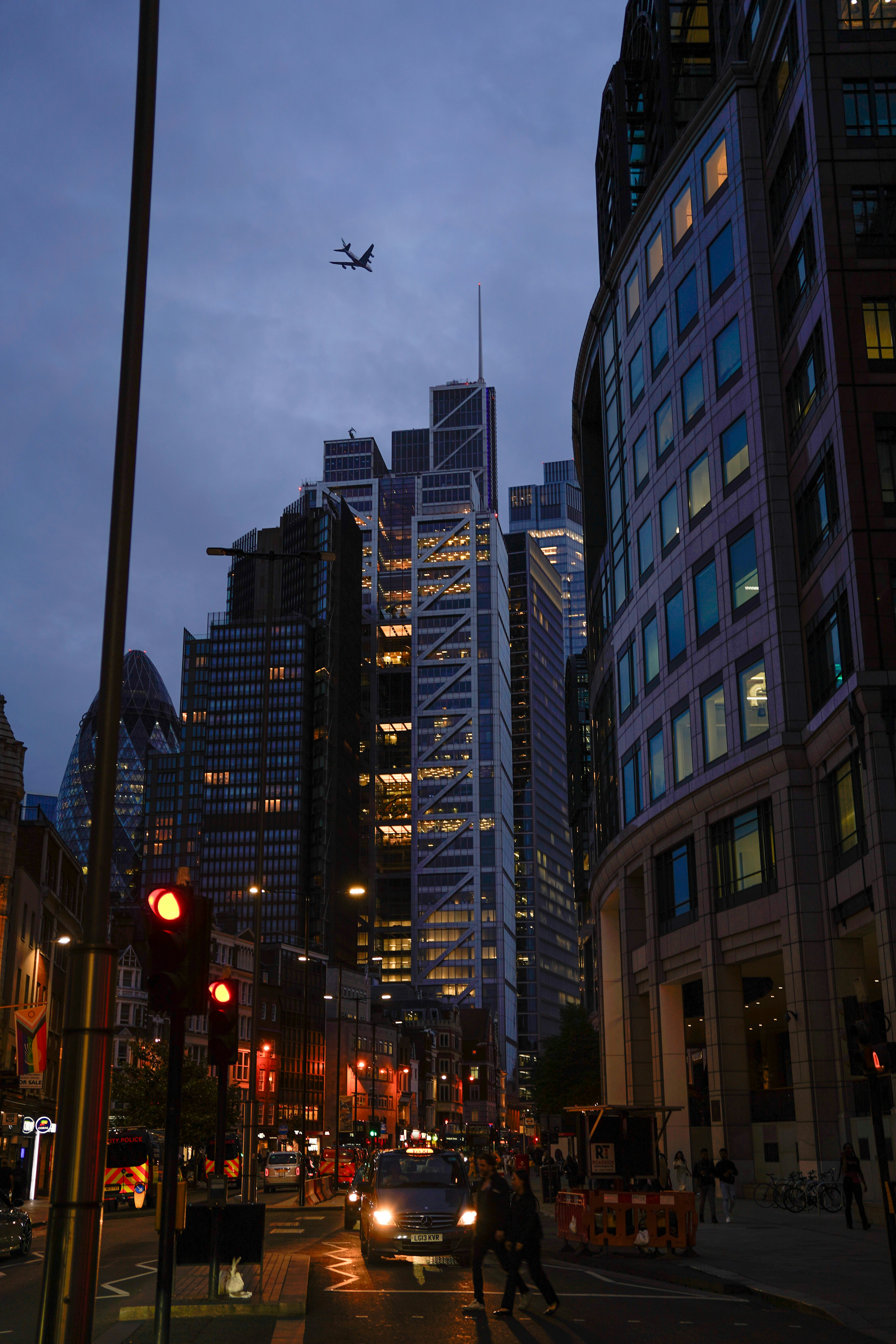 An airplane flies over illuminated city buildings at dusk.