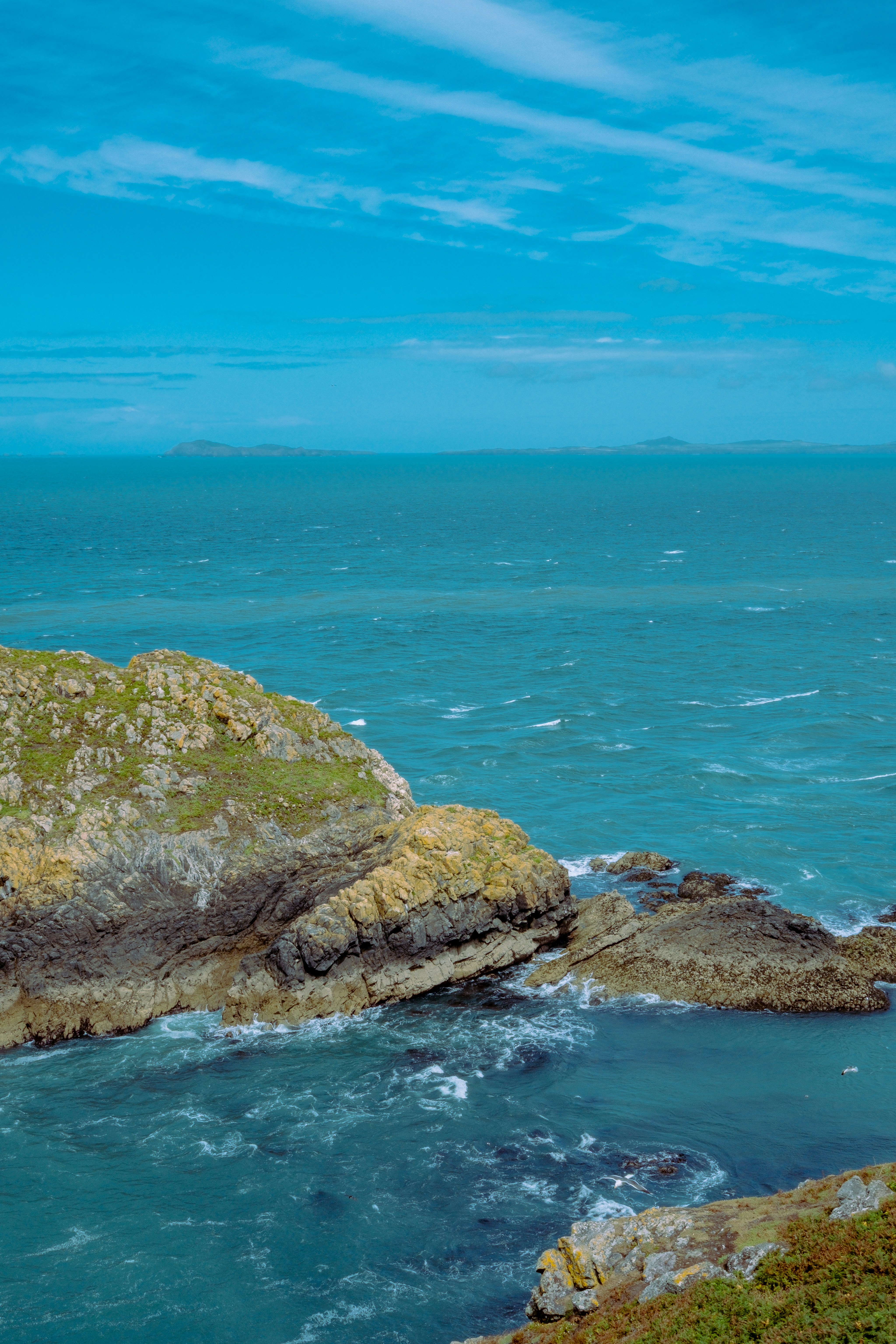 An elevated view of the rugged Pembrokeshire coastline, showcasing the striking contrast between dark cliffs and vibrant turquoise waters. The scene captures the raw beauty of the Welsh coast, with distant land visible under a bright sky. | Rocky coastline meets the sea under a blue sky.
