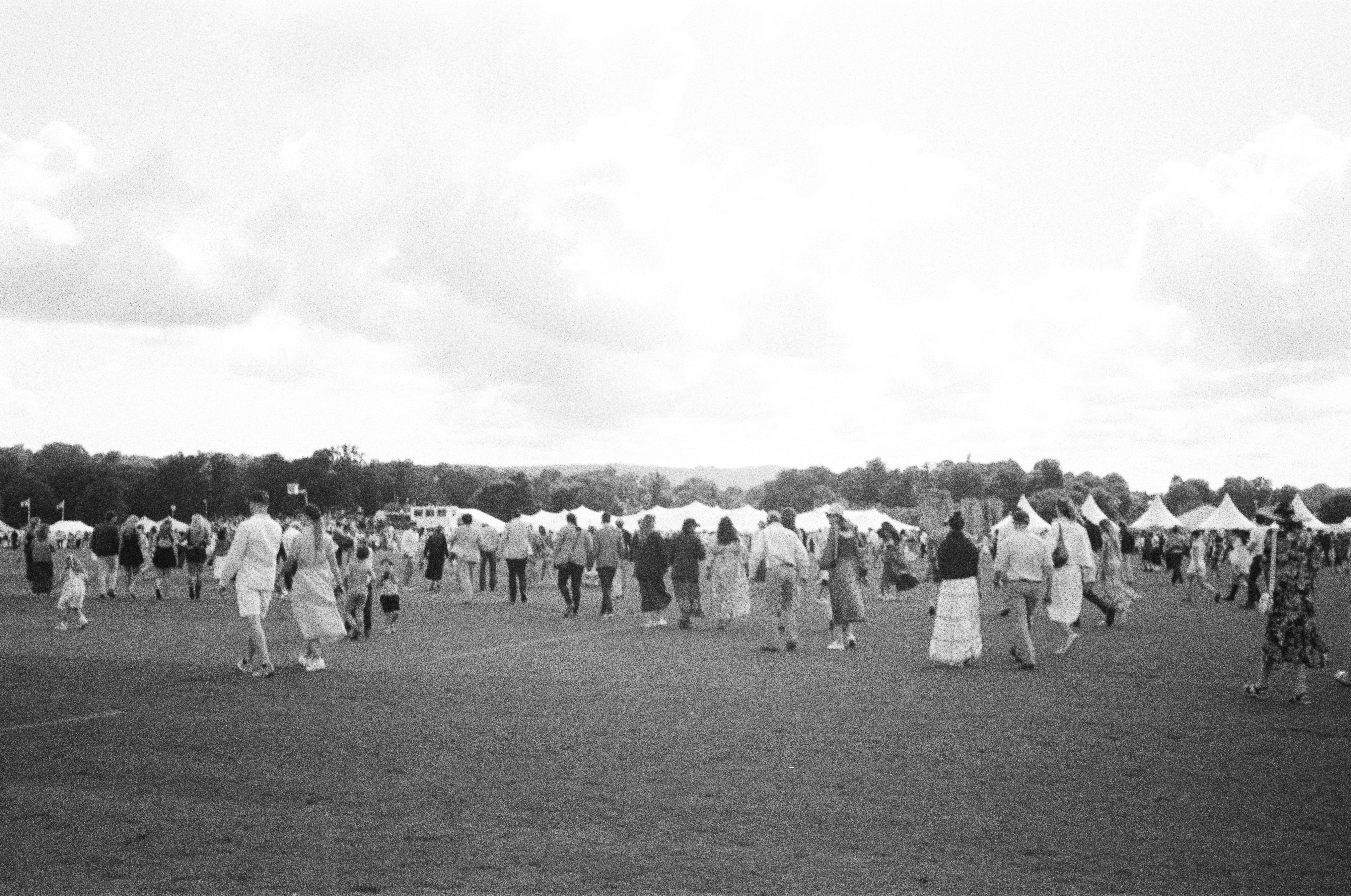 Crowd of people moving across a grassy field during an outdoor event, with tents visible in the background. The scene is captured in black and white.