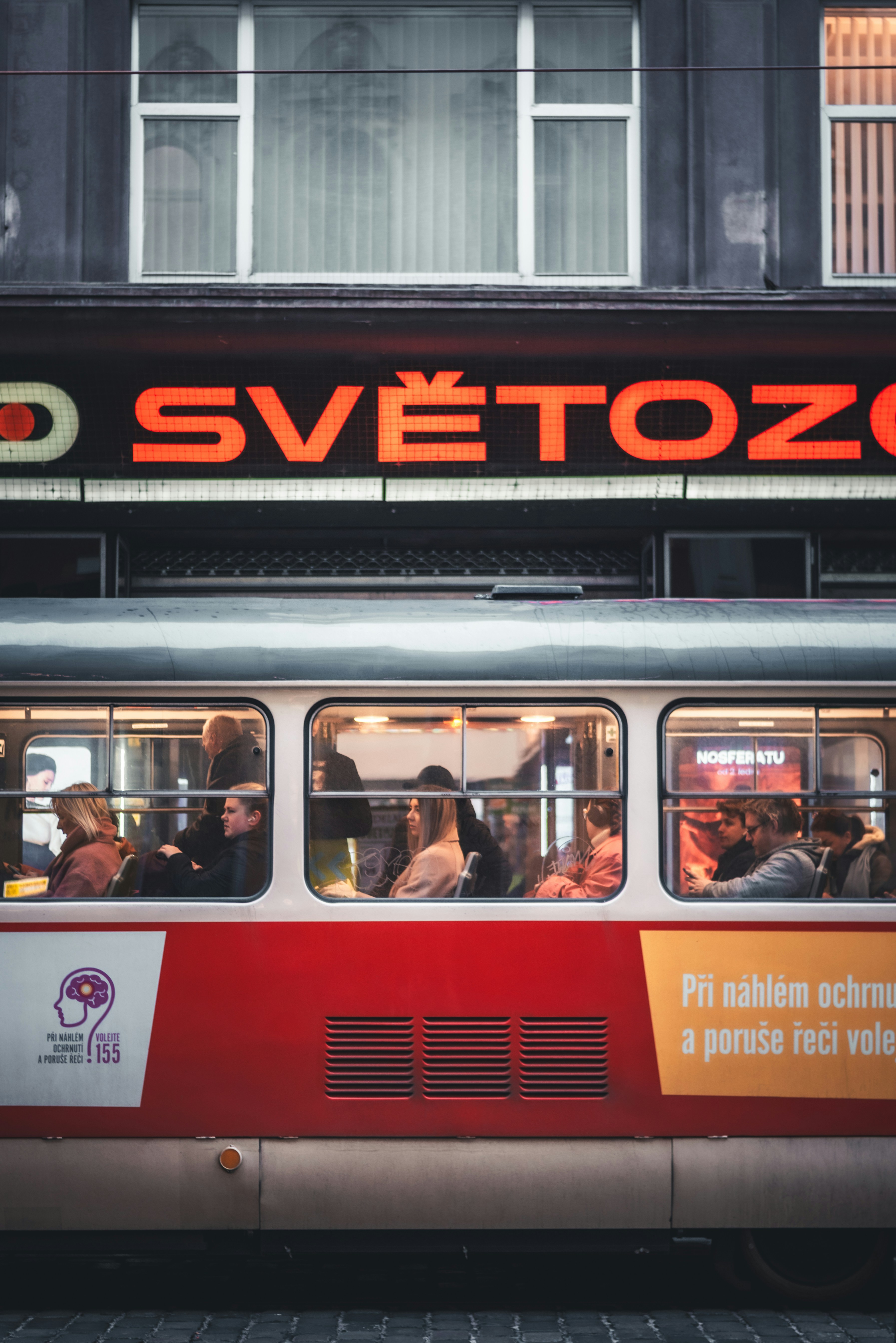 A red tram passes by a storefront with people.