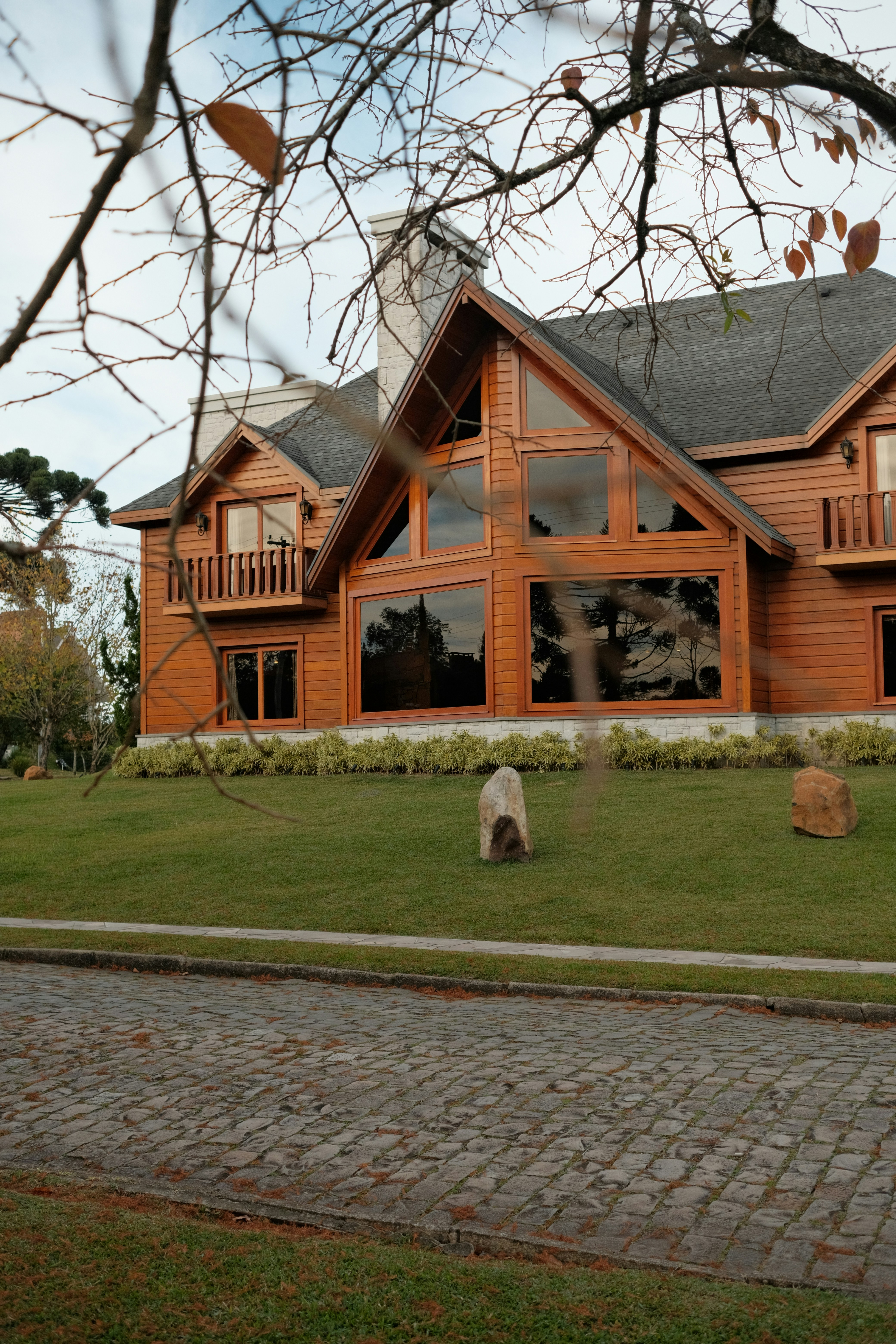 Elegant wooden house with large windows surrounded by lush greenery and a cobblestone pathway.