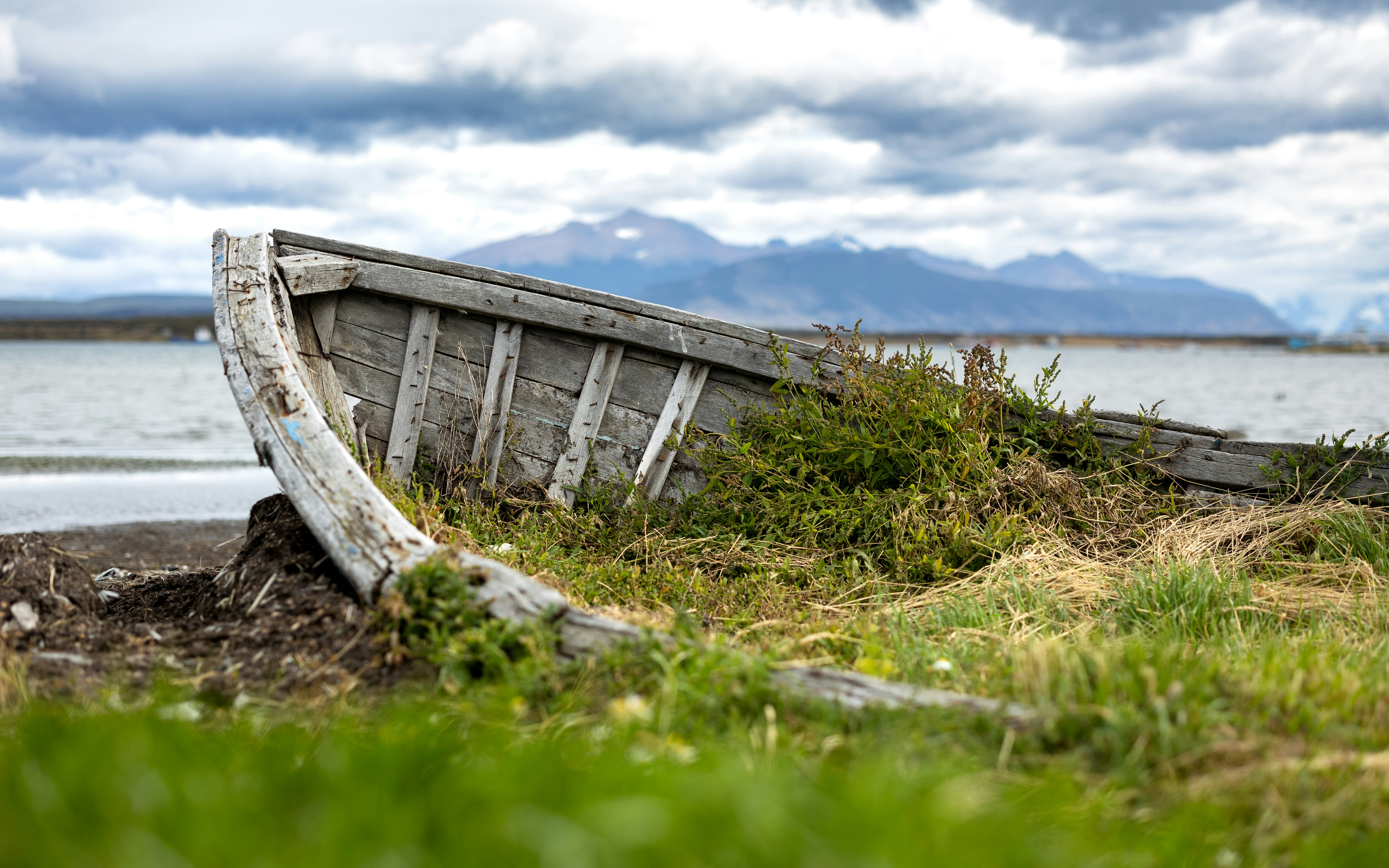 An abandoned wooden boat partially submerged in grass and sand, with mountains in the background under a cloudy sky.
