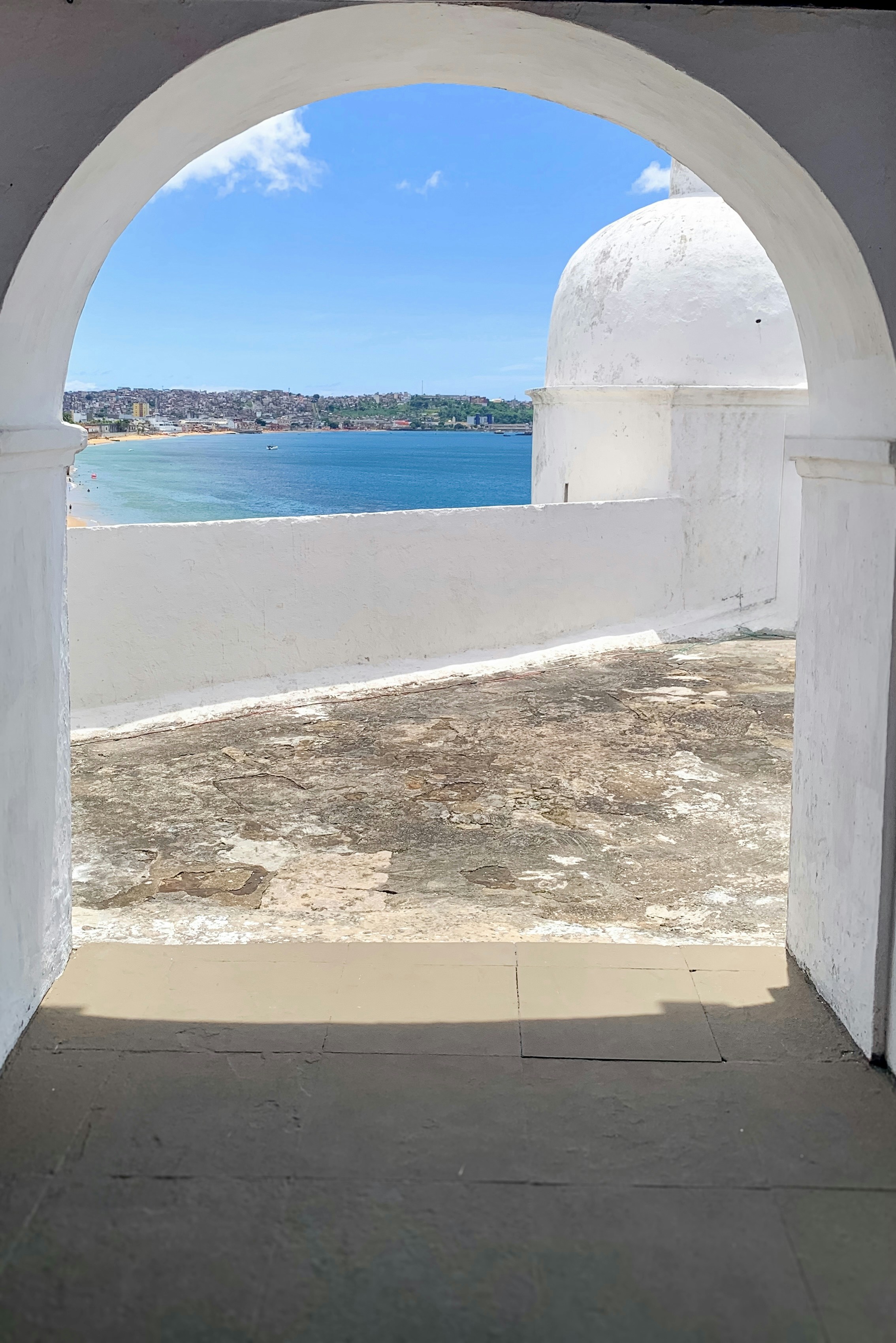 Forte de Santo Antônio da Barra, mais conhecido como o Farol da Barra, em Salvador, Bahia. Esse forte é um dos mais antigos do Brasil, originalmente construído em 1534 e reformado em 1698. Hoje abriga o Museu Náutico da Bahia e é um dos pontos turísticos mais icônicos da cidade, oferecendo uma vista privilegiada da Baía de Todos-os-Santos. O domo branco visível na imagem é uma característica marcante da arquitetura do forte. | Arched doorway frames a view of the sea.