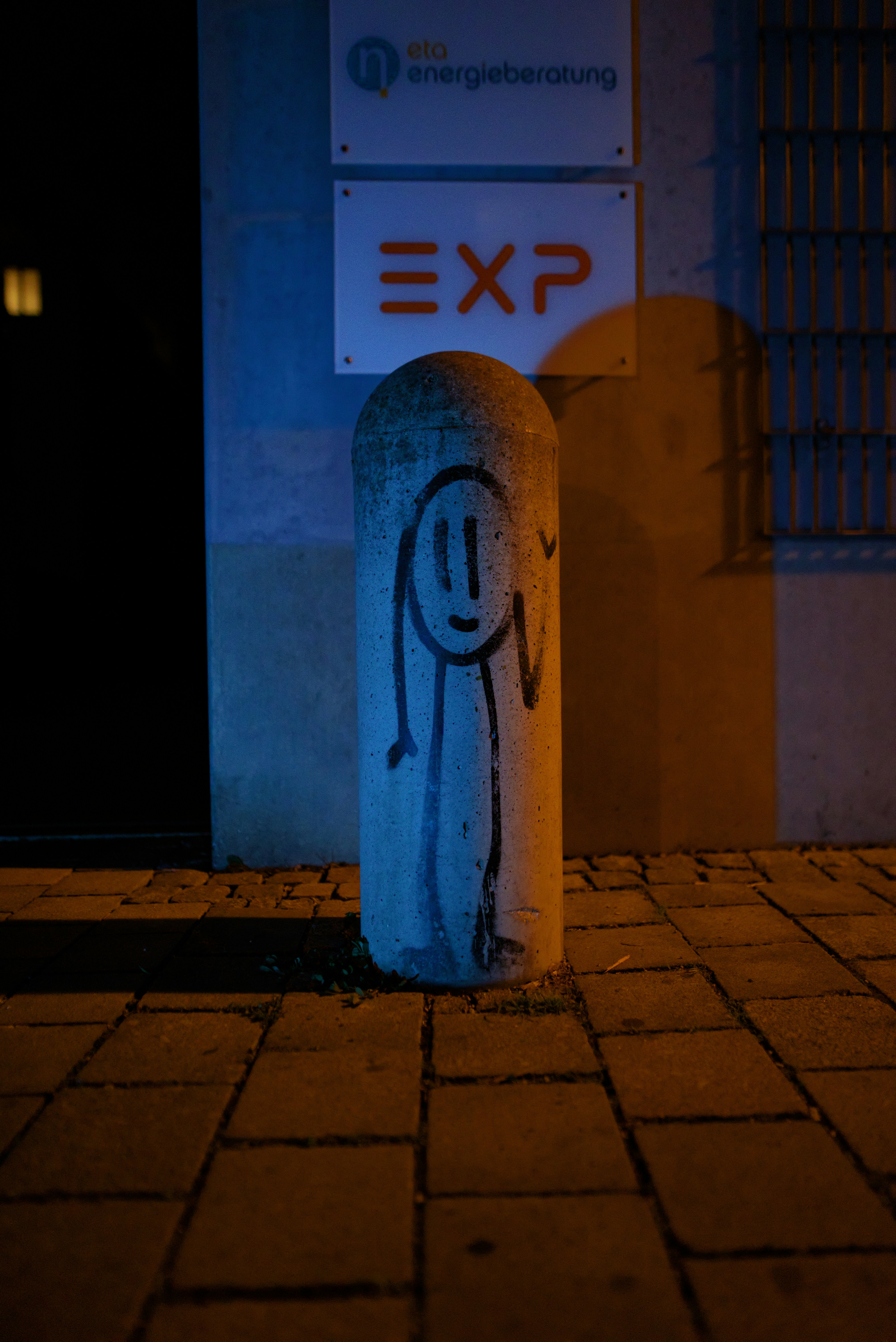 A playful graffiti figure on a concrete post stands against a softly lit urban backdrop, showcasing the intersection of art and street life.