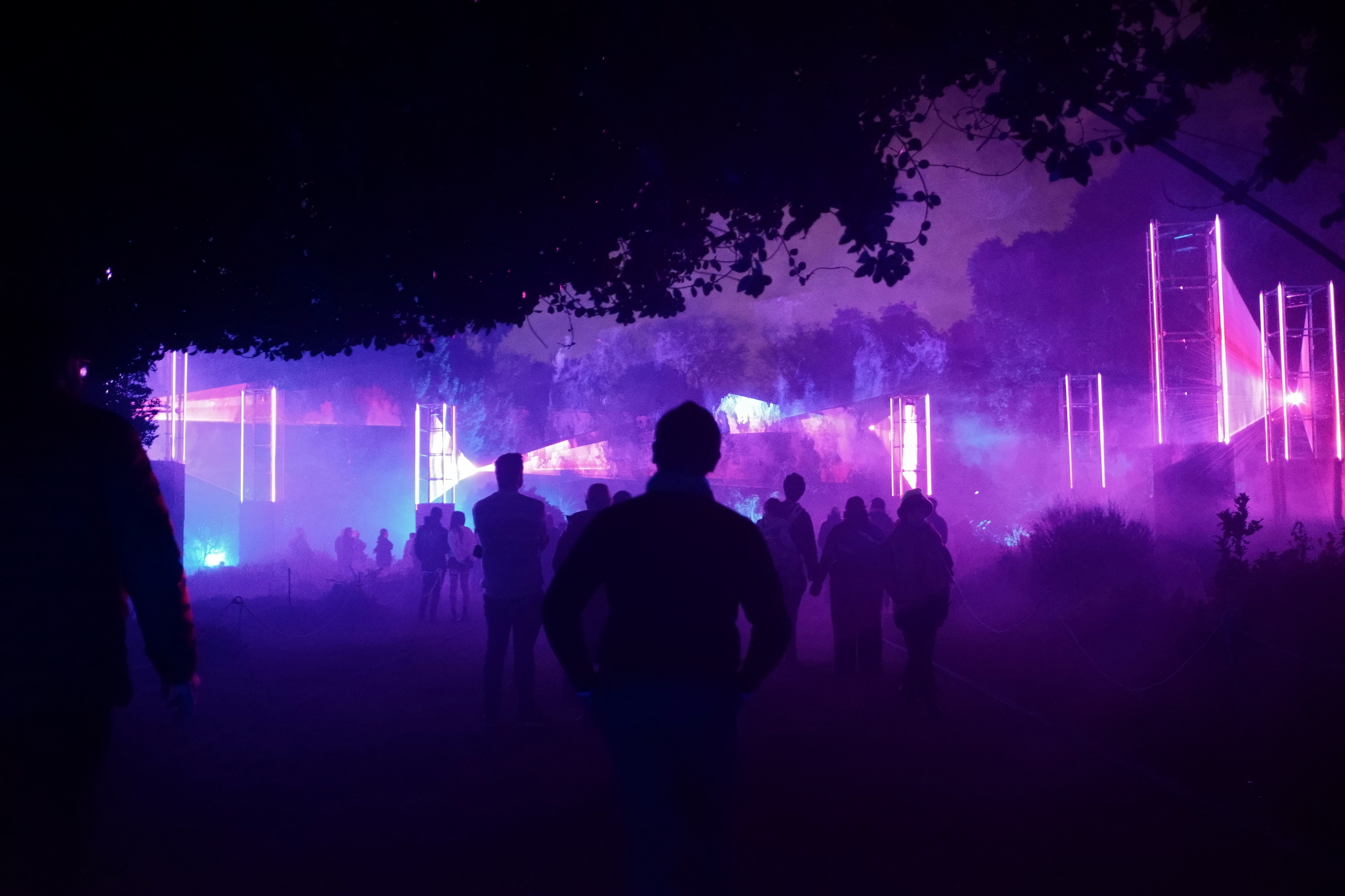 Silhouetted figures wander through a vibrant, fog-filled landscape illuminated by colorful lights and neon structures.